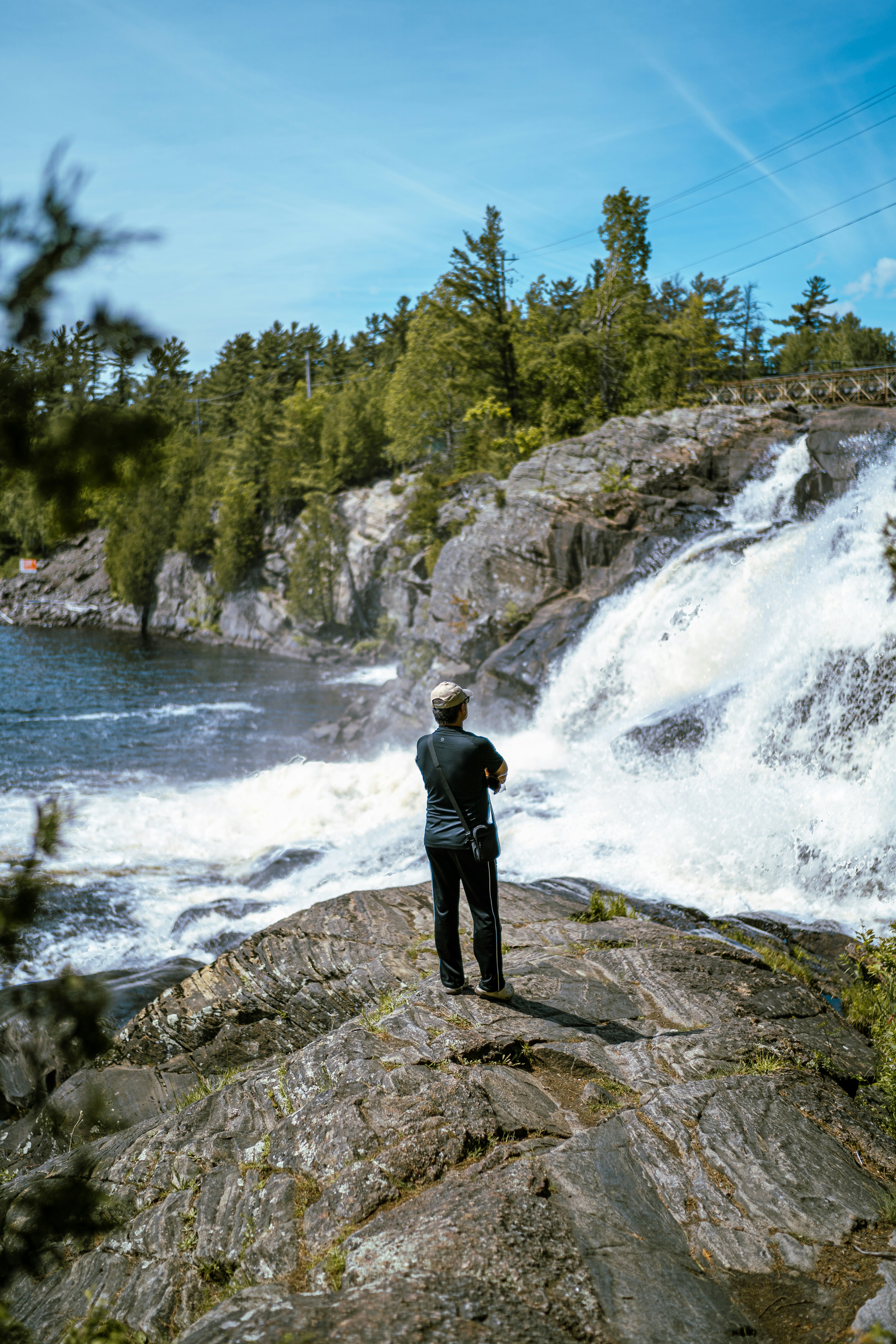 A lone figure stands on rocky terrain, gazing at a powerful waterfall surrounded by lush greenery. The scene captures the essence of tranquility and the awe of nature.