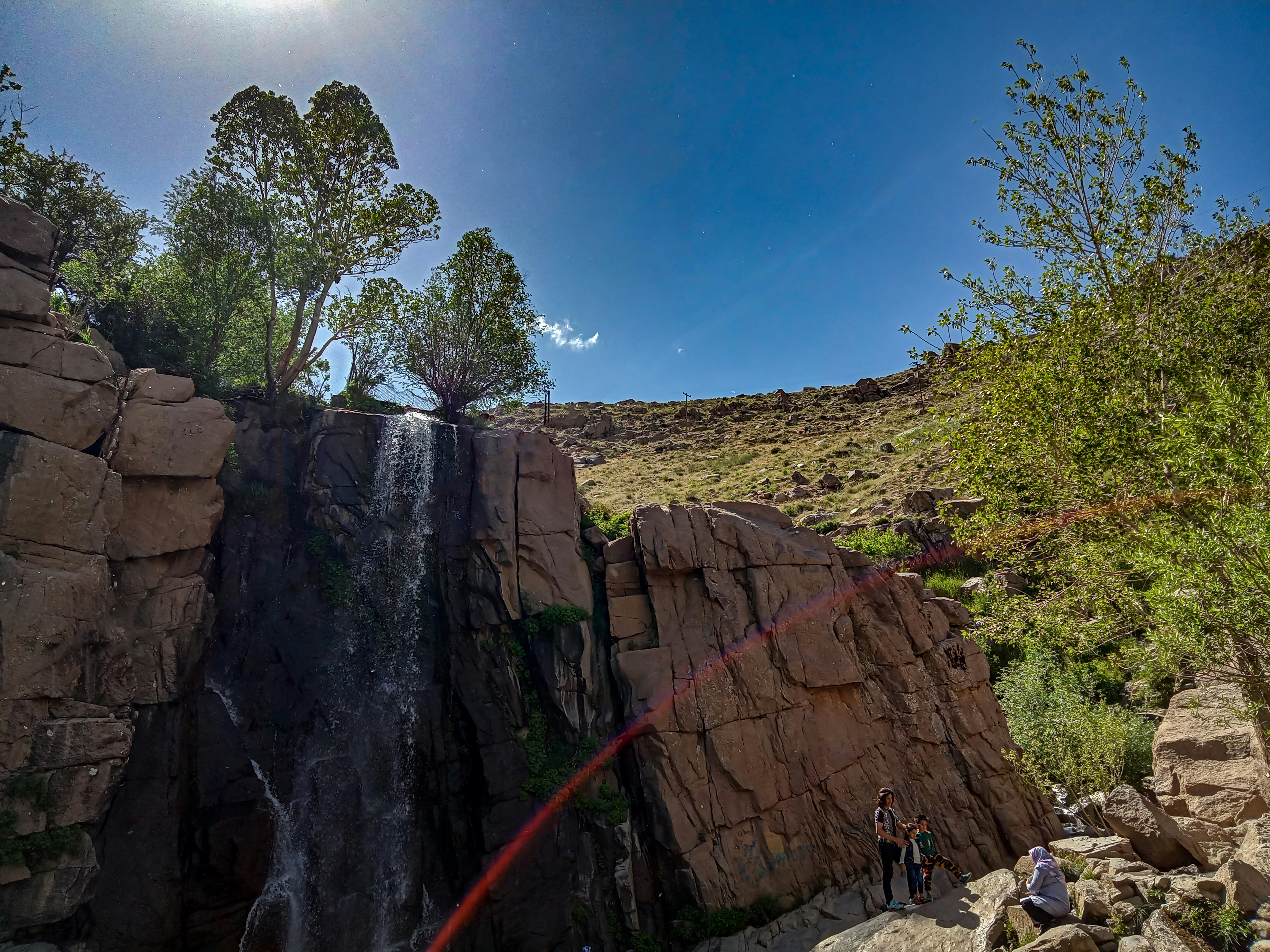 Waterfall cascading down rugged cliffs under a bright sky, surrounded by leafy trees and hikers exploring the terrain.