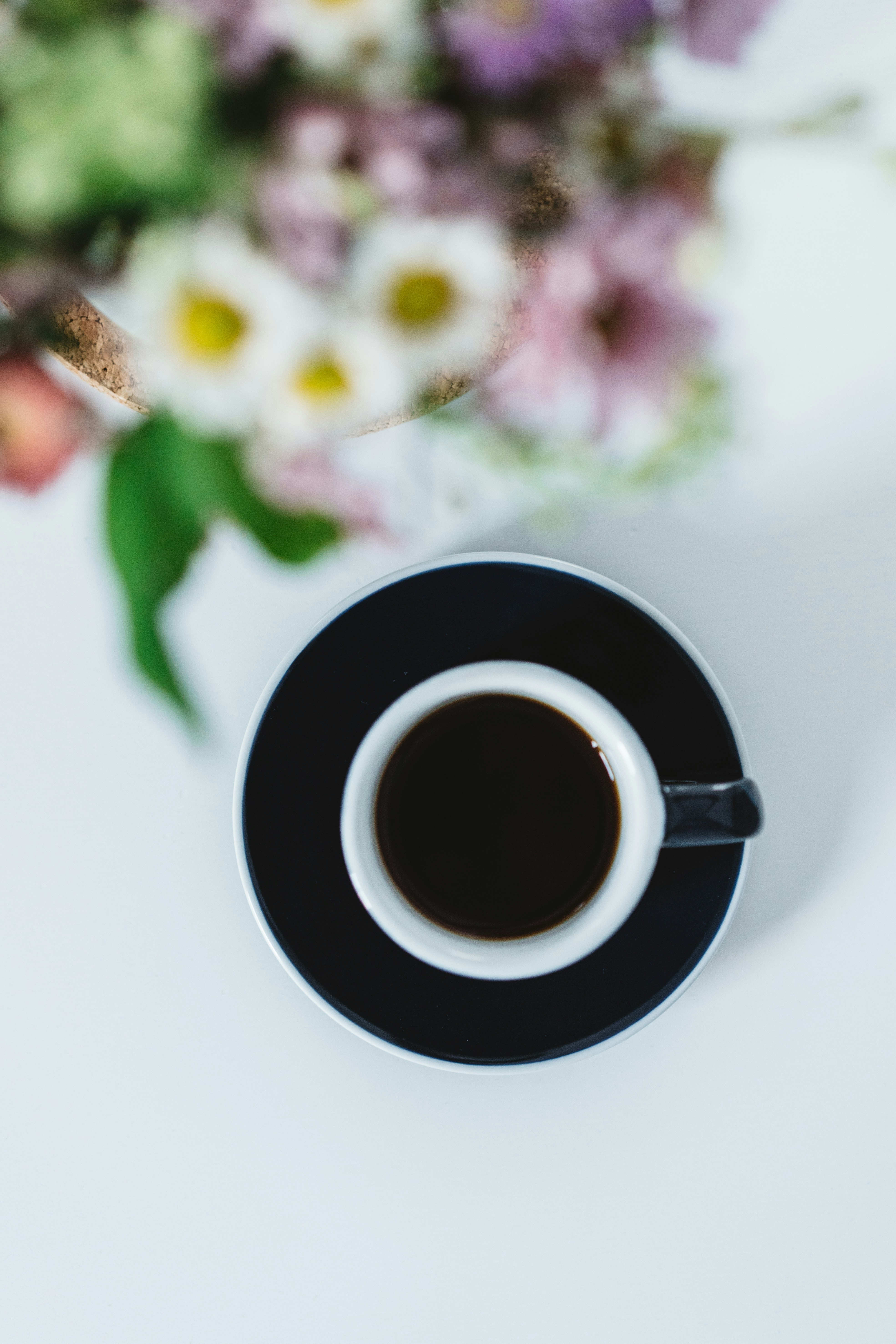 A black coffee cup sits on a white surface, surrounded by softly blurred flowers in the background.