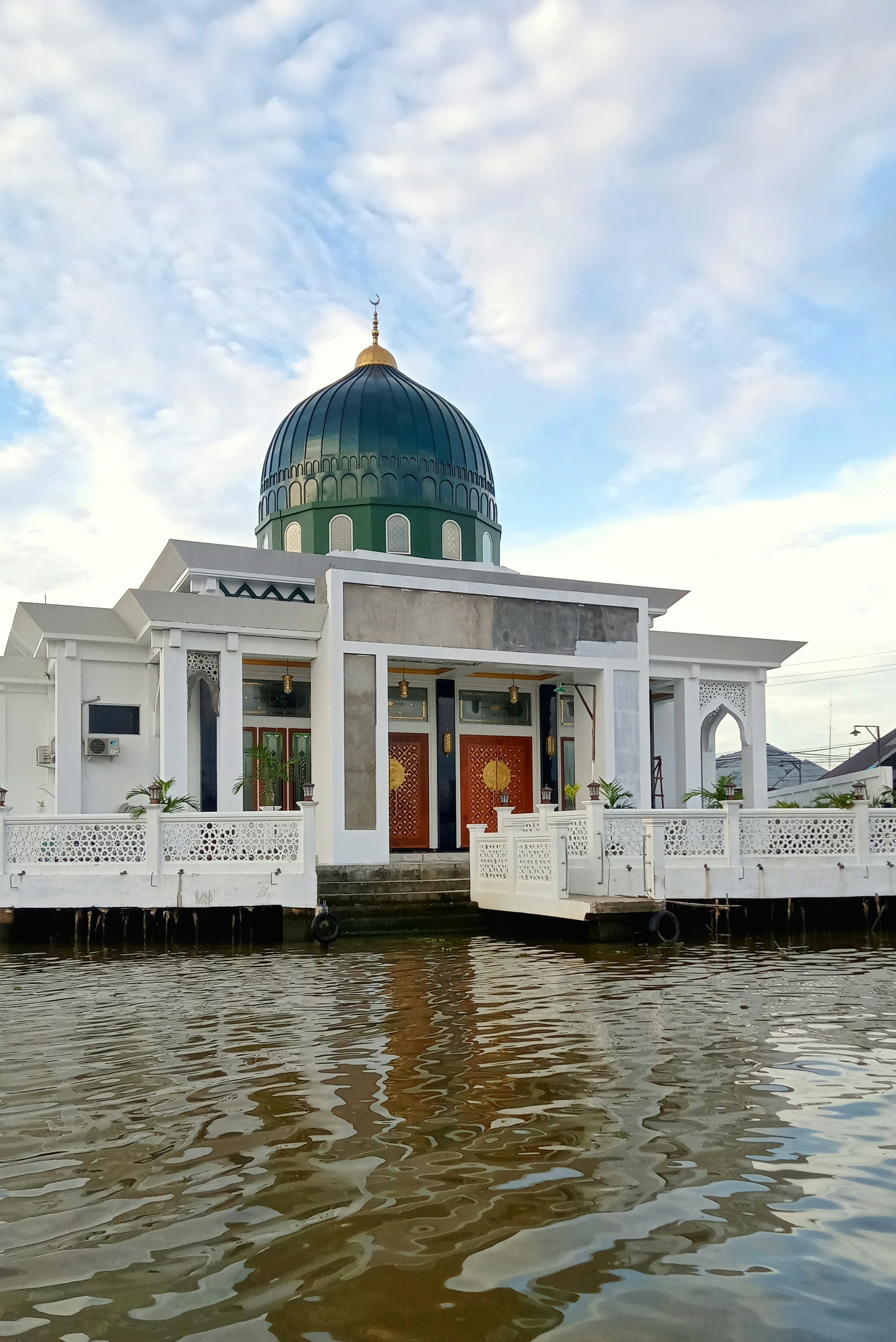 A beautifully designed mosque with a green dome, situated on the edge of a tranquil body of water, surrounded by decorative white railings. The sky is partly cloudy, enhancing the serene atmosphere.
