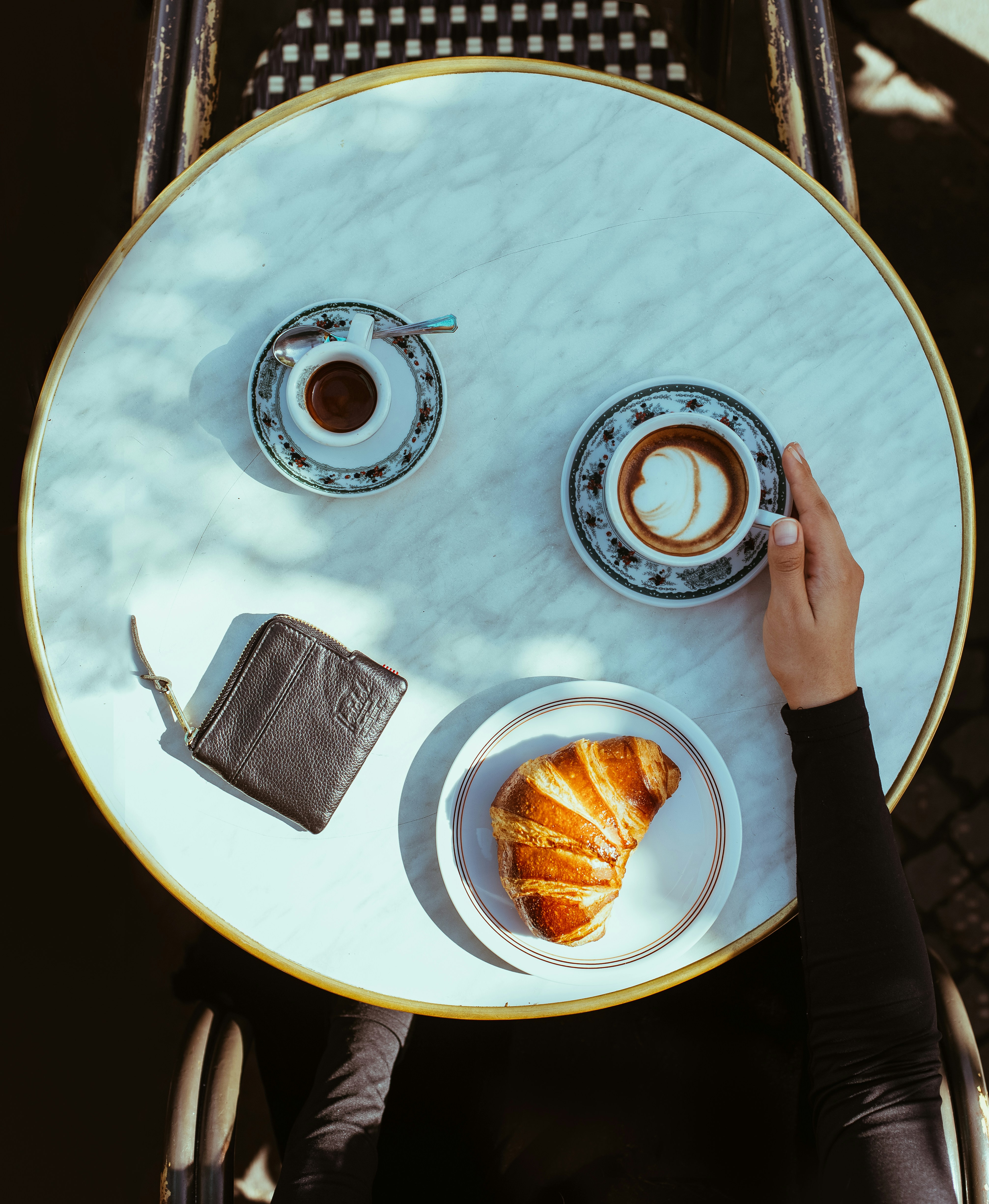 A beautifully arranged breakfast scene featuring a croissant, coffee, and a wallet on a marble table. The warm light enhances the inviting atmosphere.