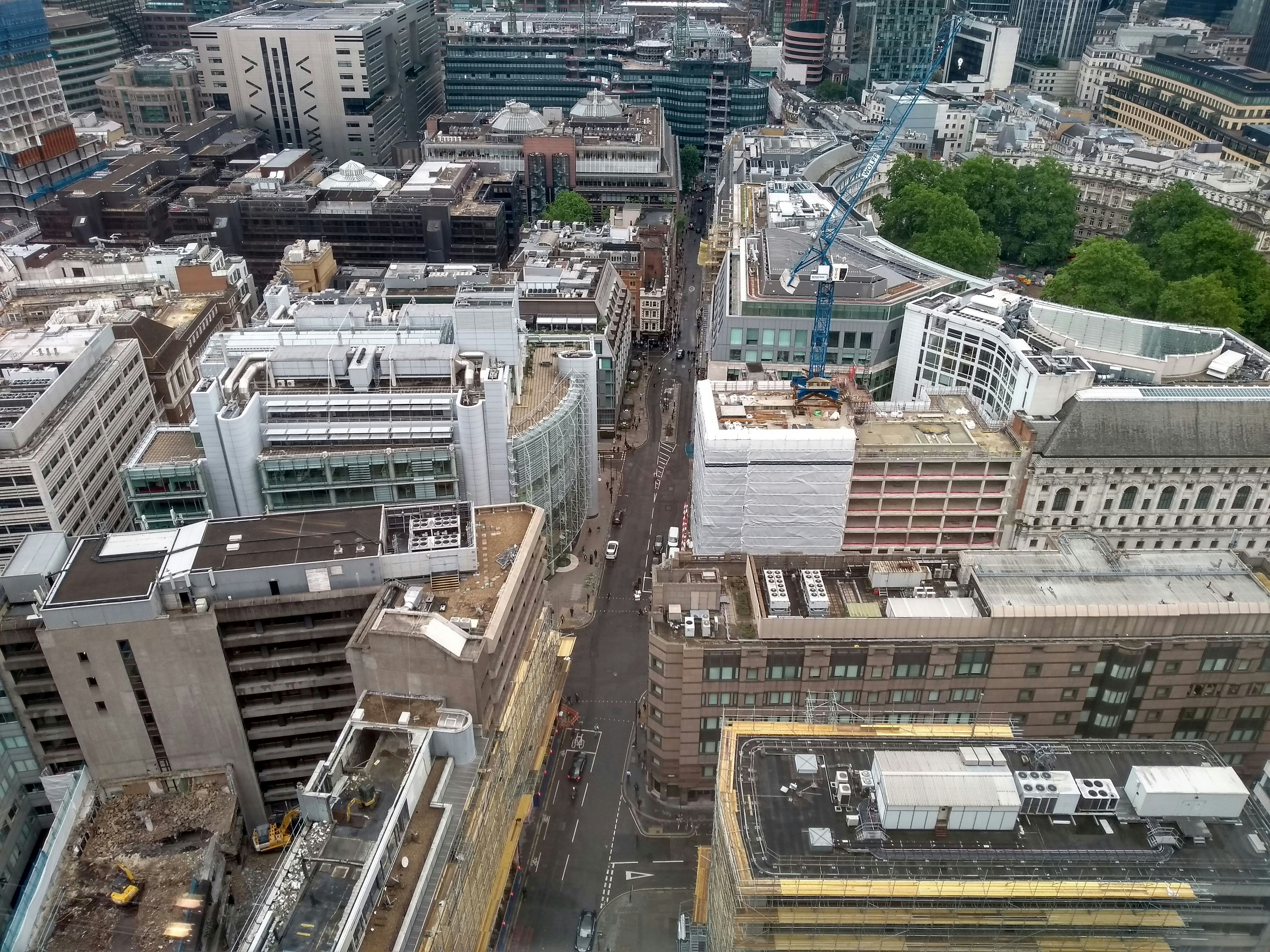 Aerial photograph of a dense urban block with a central street cutting through grey and brown buildings. A construction crane and pockets of green trees punctuate the skyline.