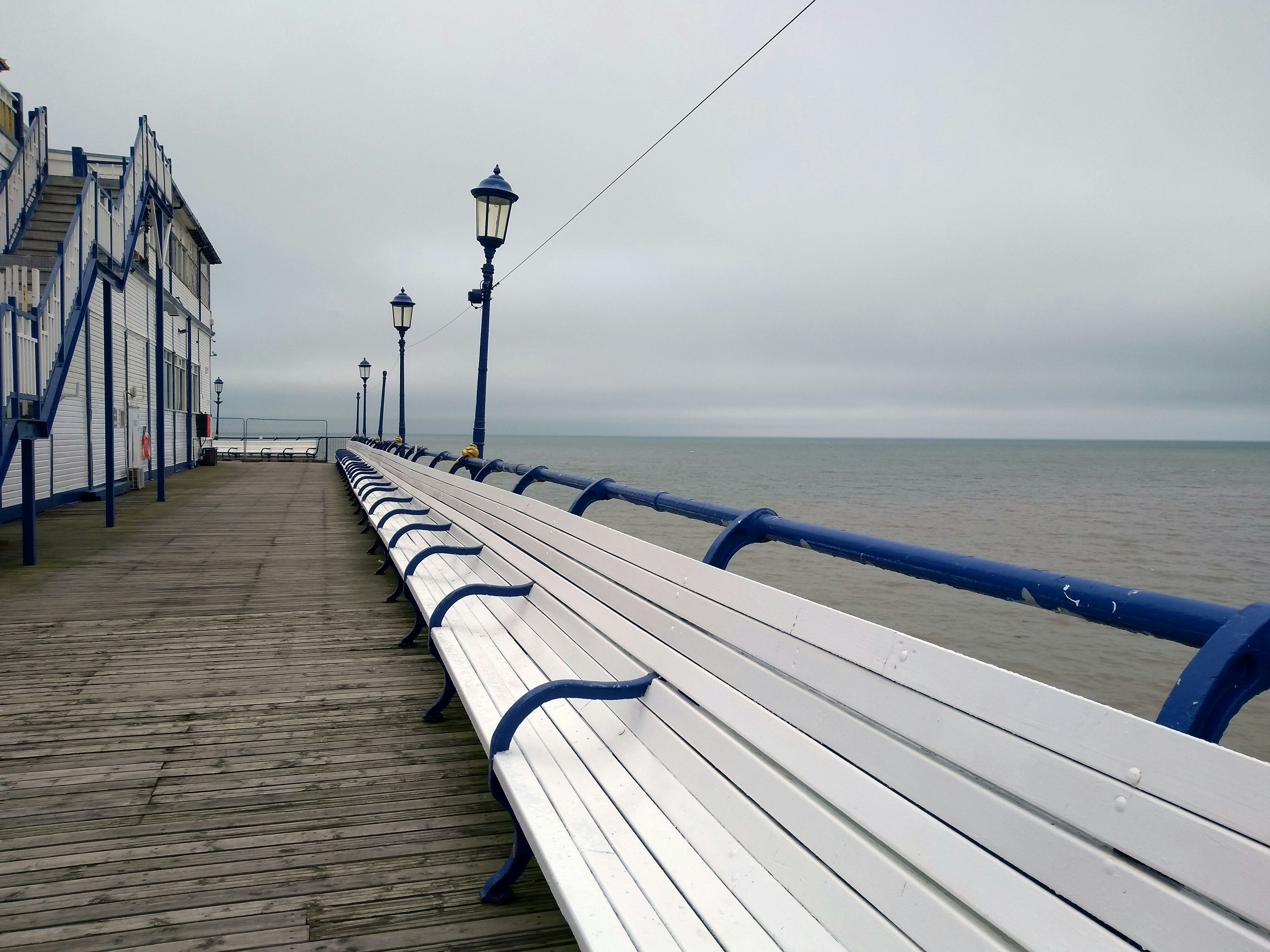 Row of white benches lines the wooden pier, guiding the eye toward the calm sea at the horizon. Overcast skies wash the scene in soft, muted tones.