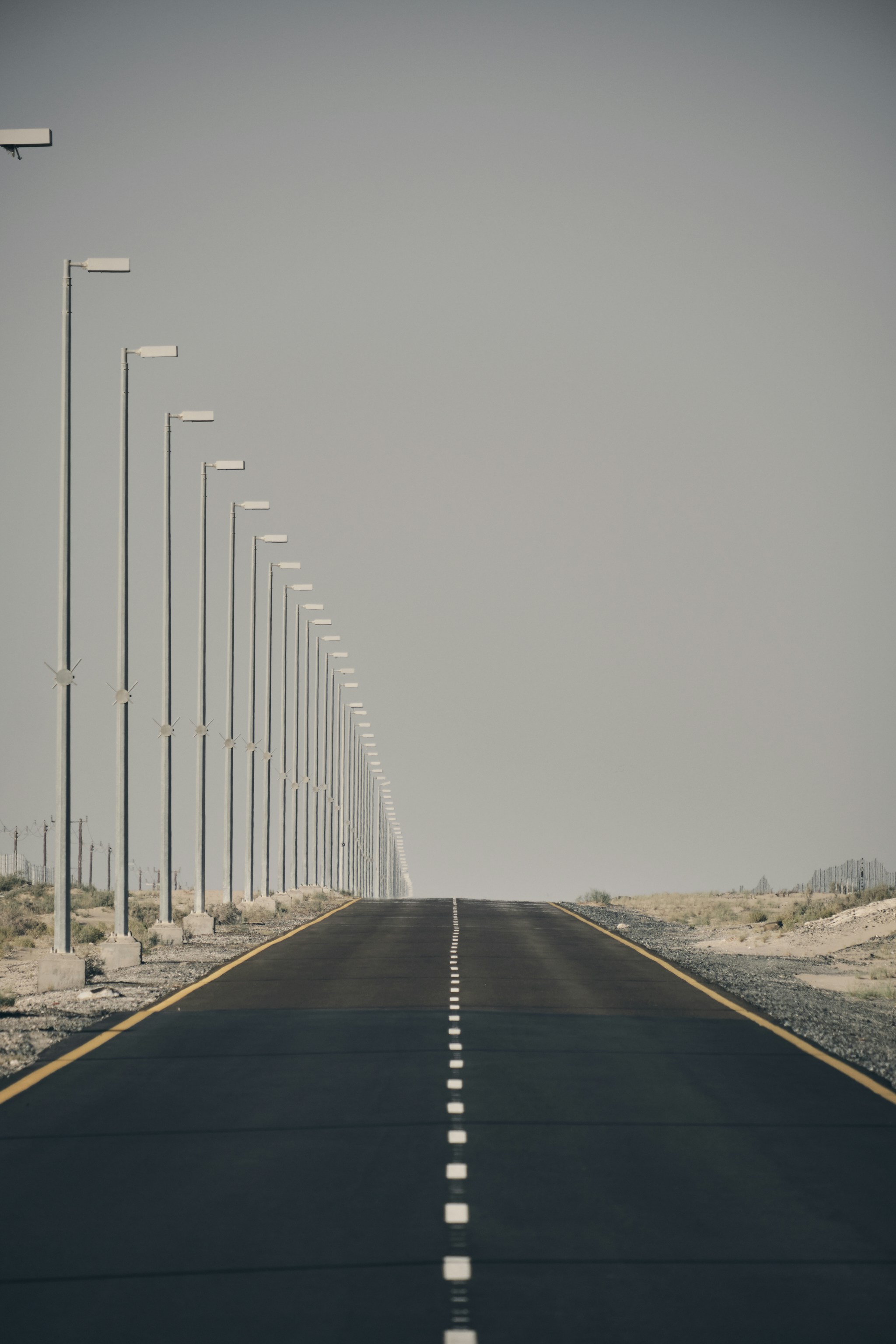 A long, straight road flanked by evenly spaced streetlights, leading towards a distant horizon under a clear sky.