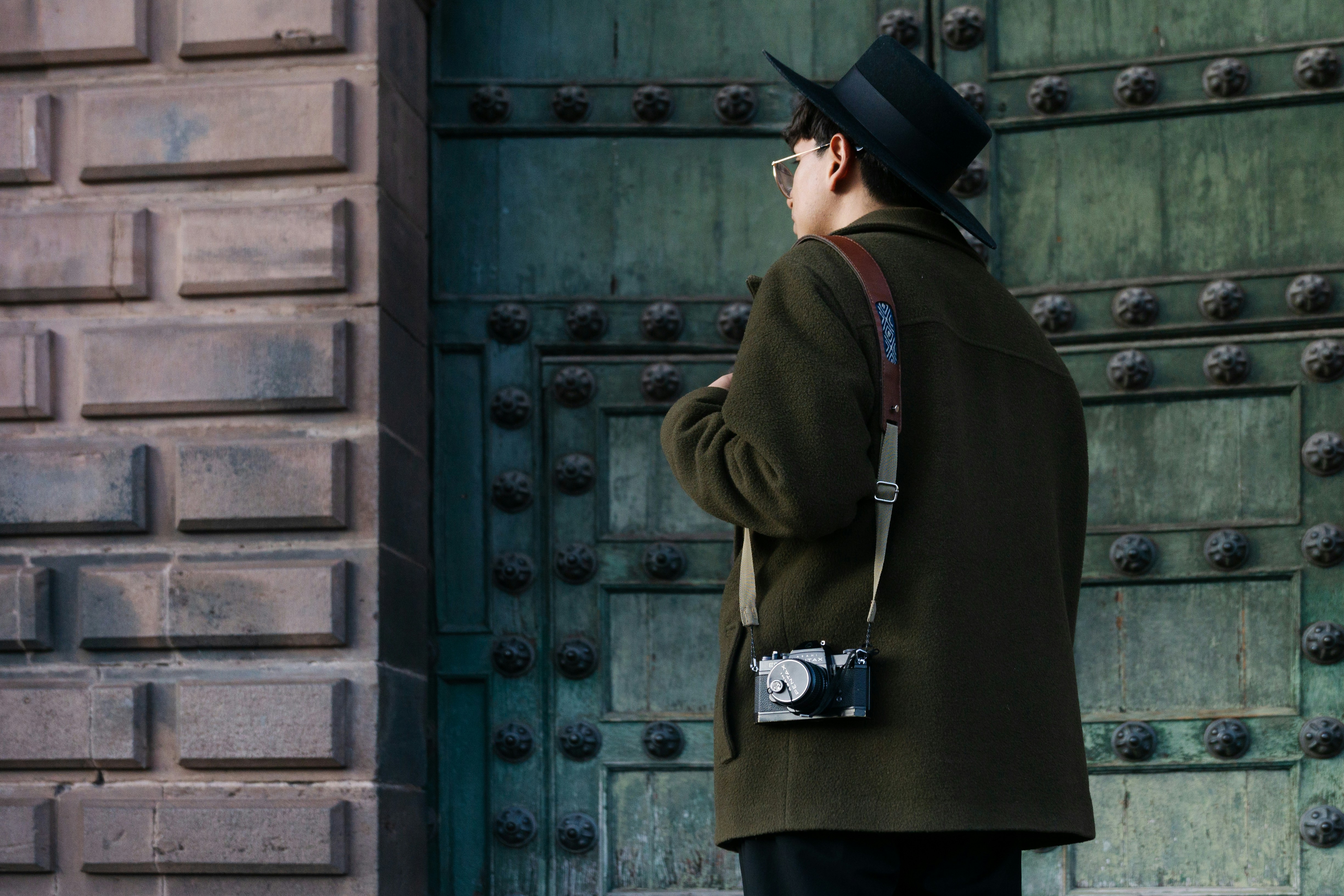 A person dressed in a dark coat and hat stands against an ornate green door, holding a camera, embodying a blend of modern photography and classic style.