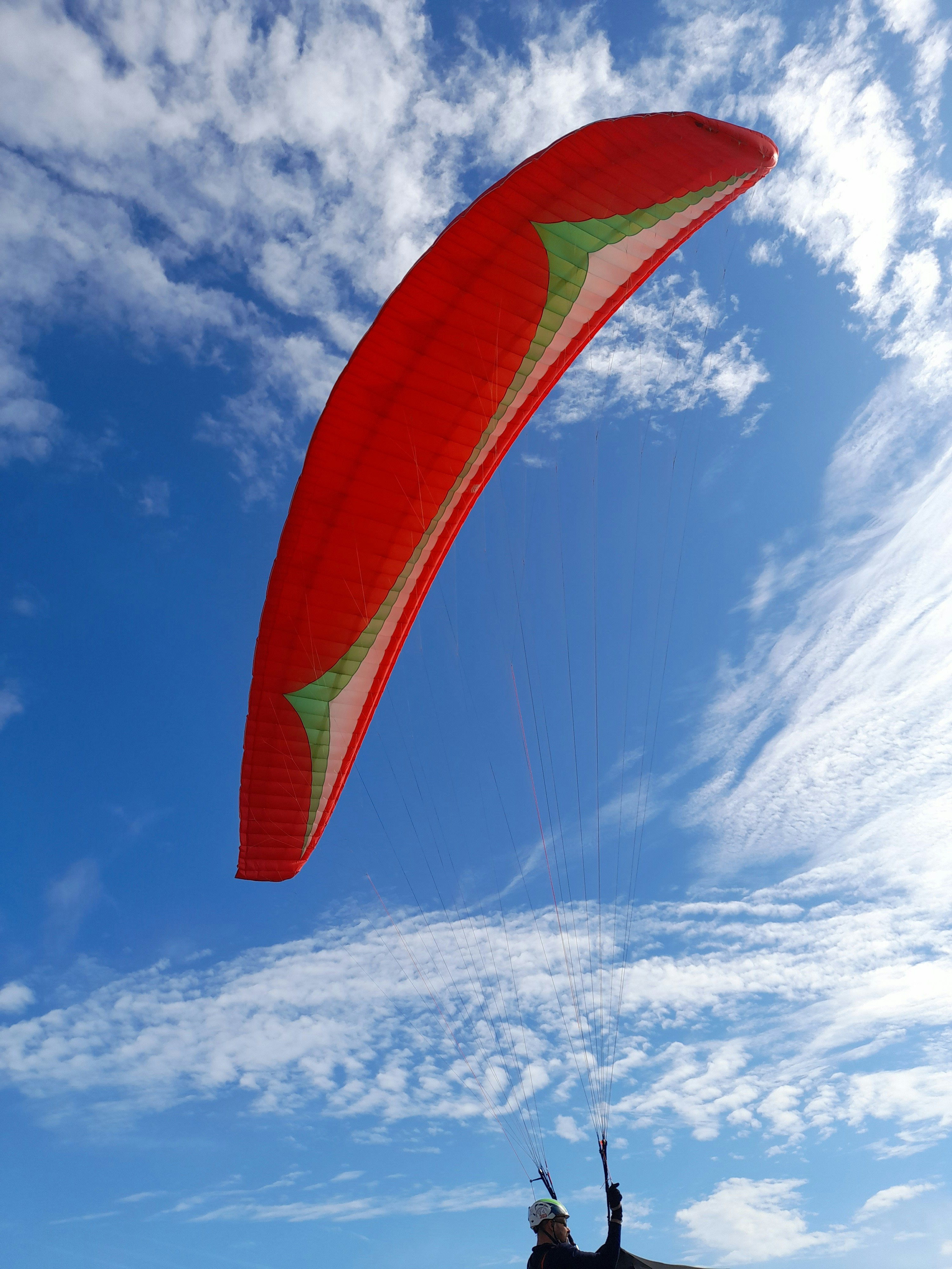A vibrant red and green paraglider is held aloft against a backdrop of blue sky and wispy clouds.