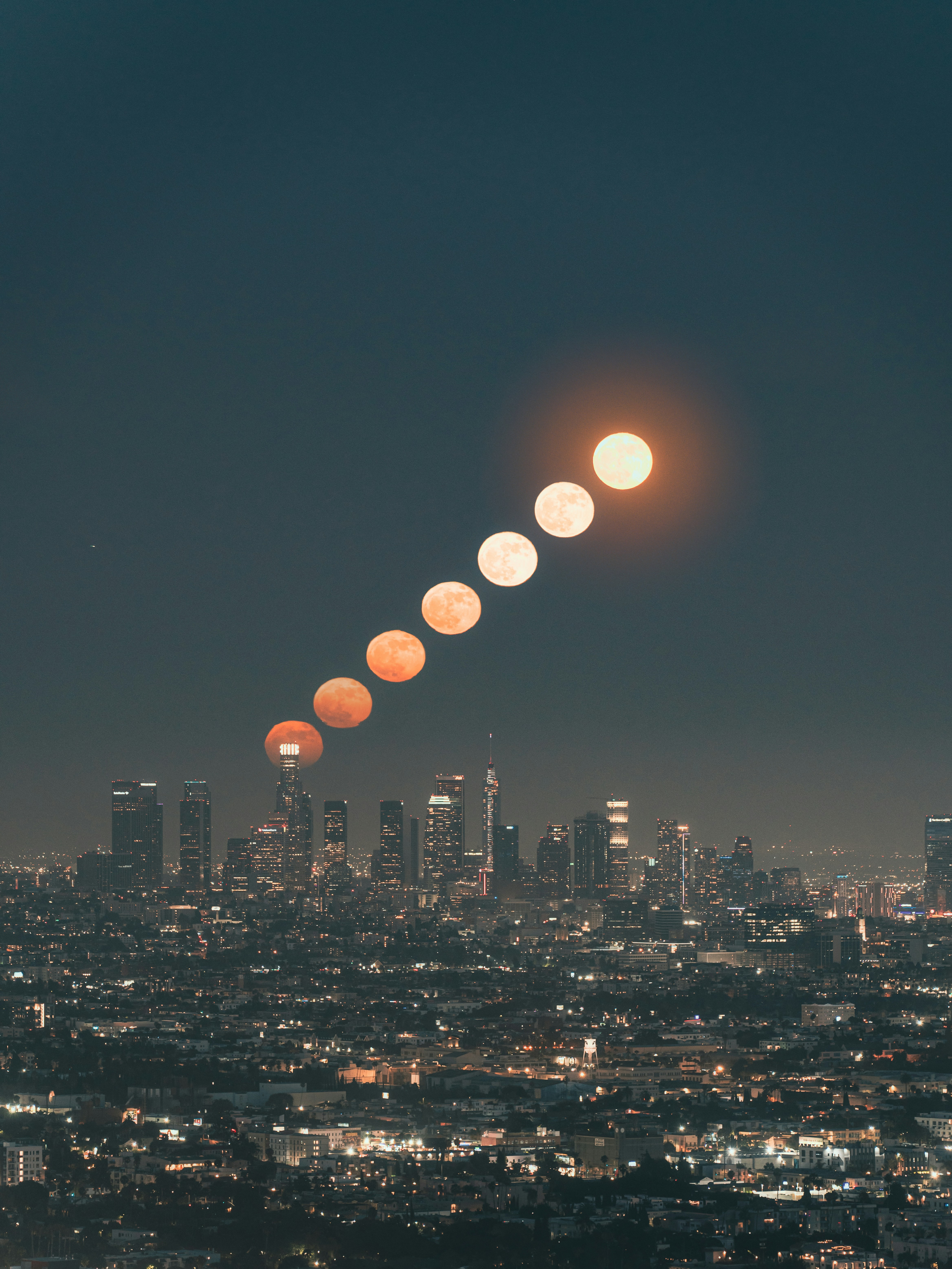 Sequence of moon phases rising over a sprawling city skyline at dusk.