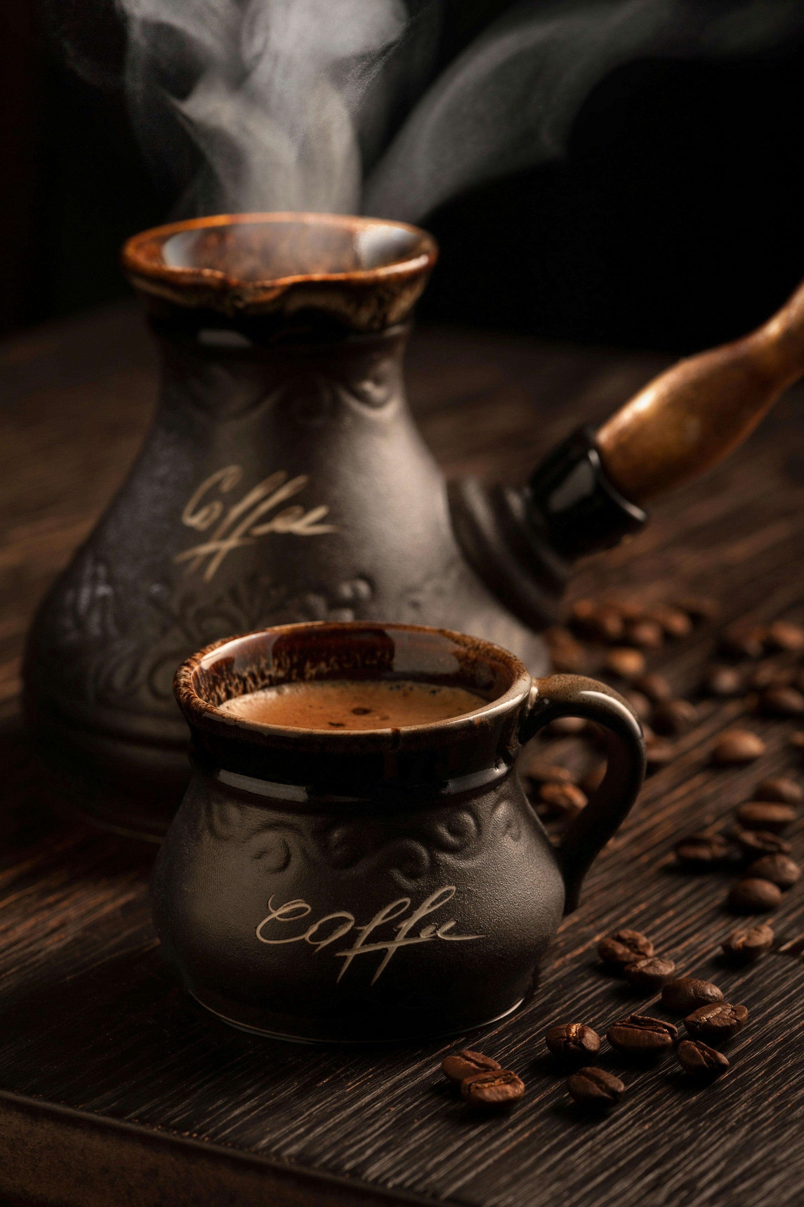 Steam rises from a traditional coffee pot alongside a small cup, both elegantly adorned with the word 'coffee.' Coffee beans scatter across a dark wooden surface.