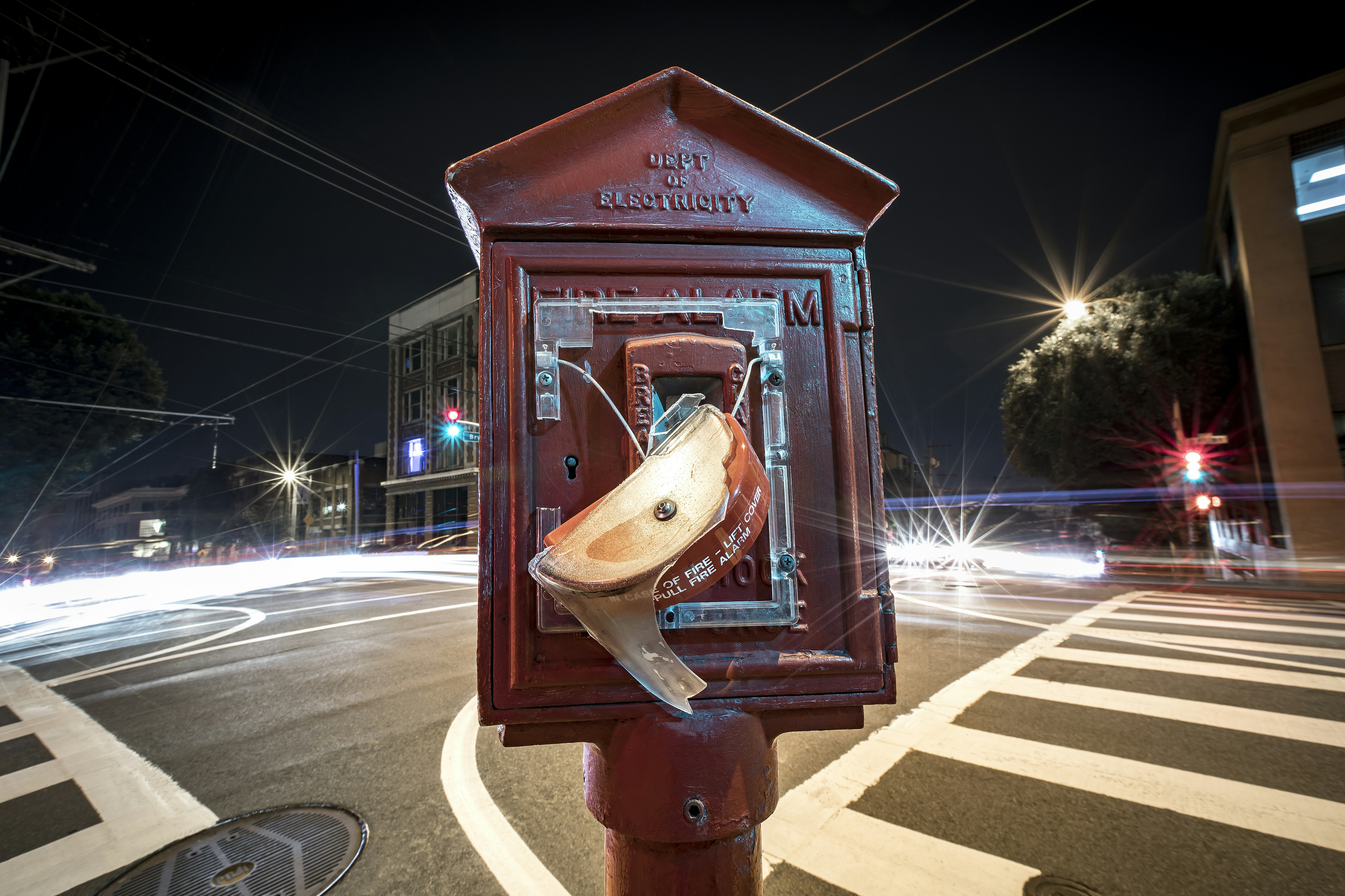 A vintage red call box with a partially detached cover stands at a city intersection, illuminated by passing vehicle lights at night.