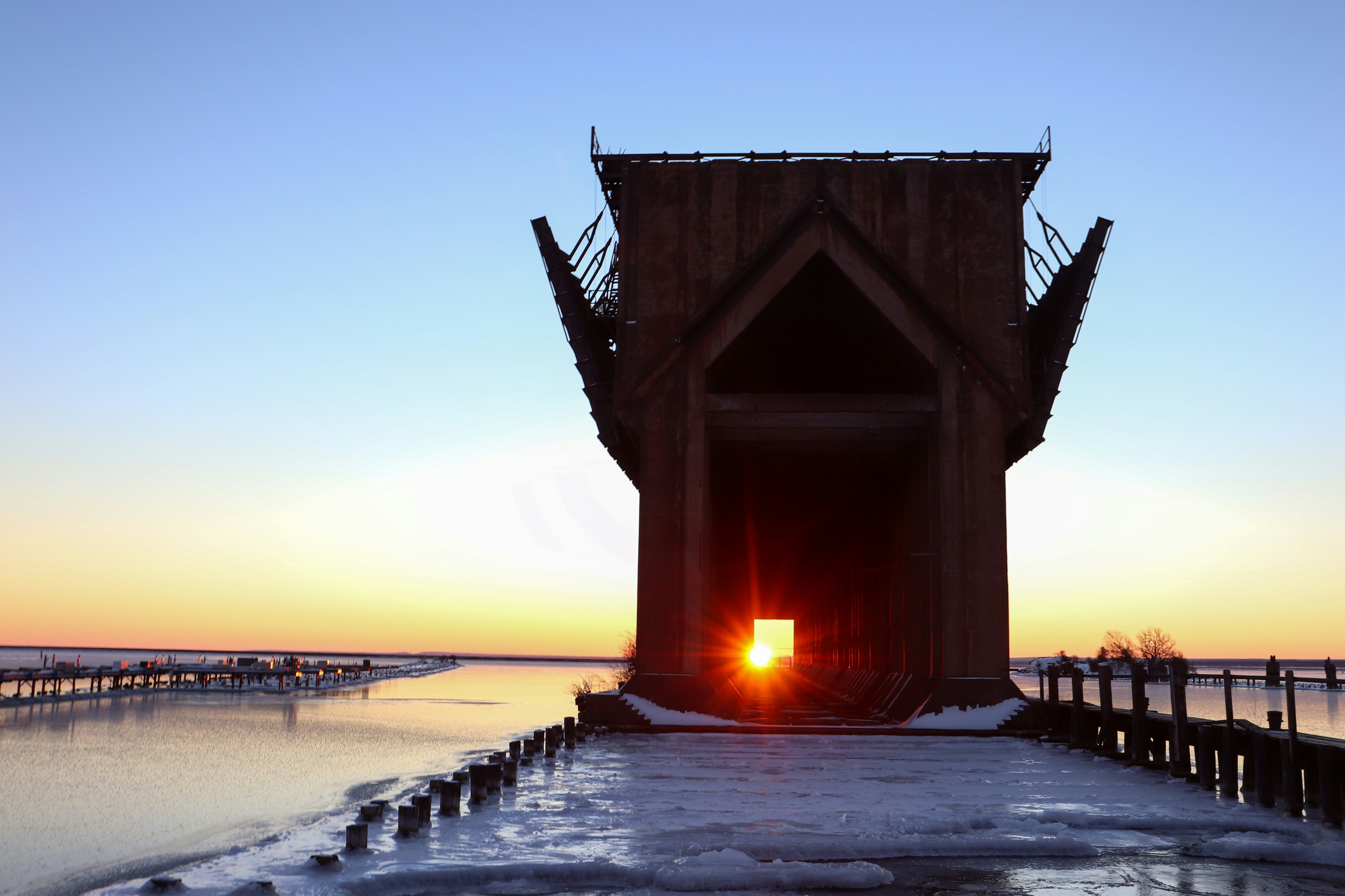 Silhouette of a large industrial structure against a vibrant sunset, with reflections on the icy water below.
