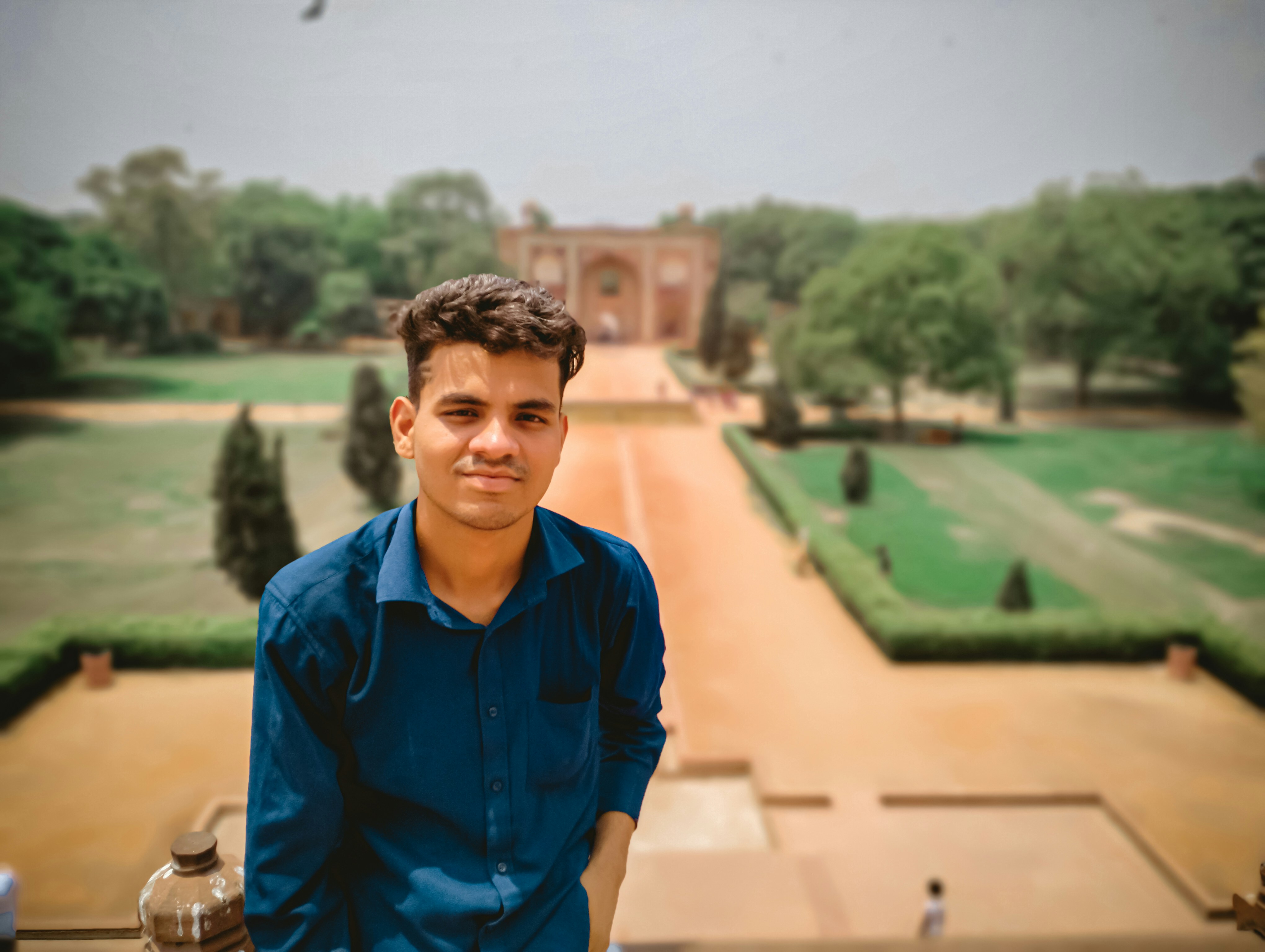Young man in a blue shirt poses in front of a historical site, surrounded by lush greenery and pathways leading to an ornate gateway.