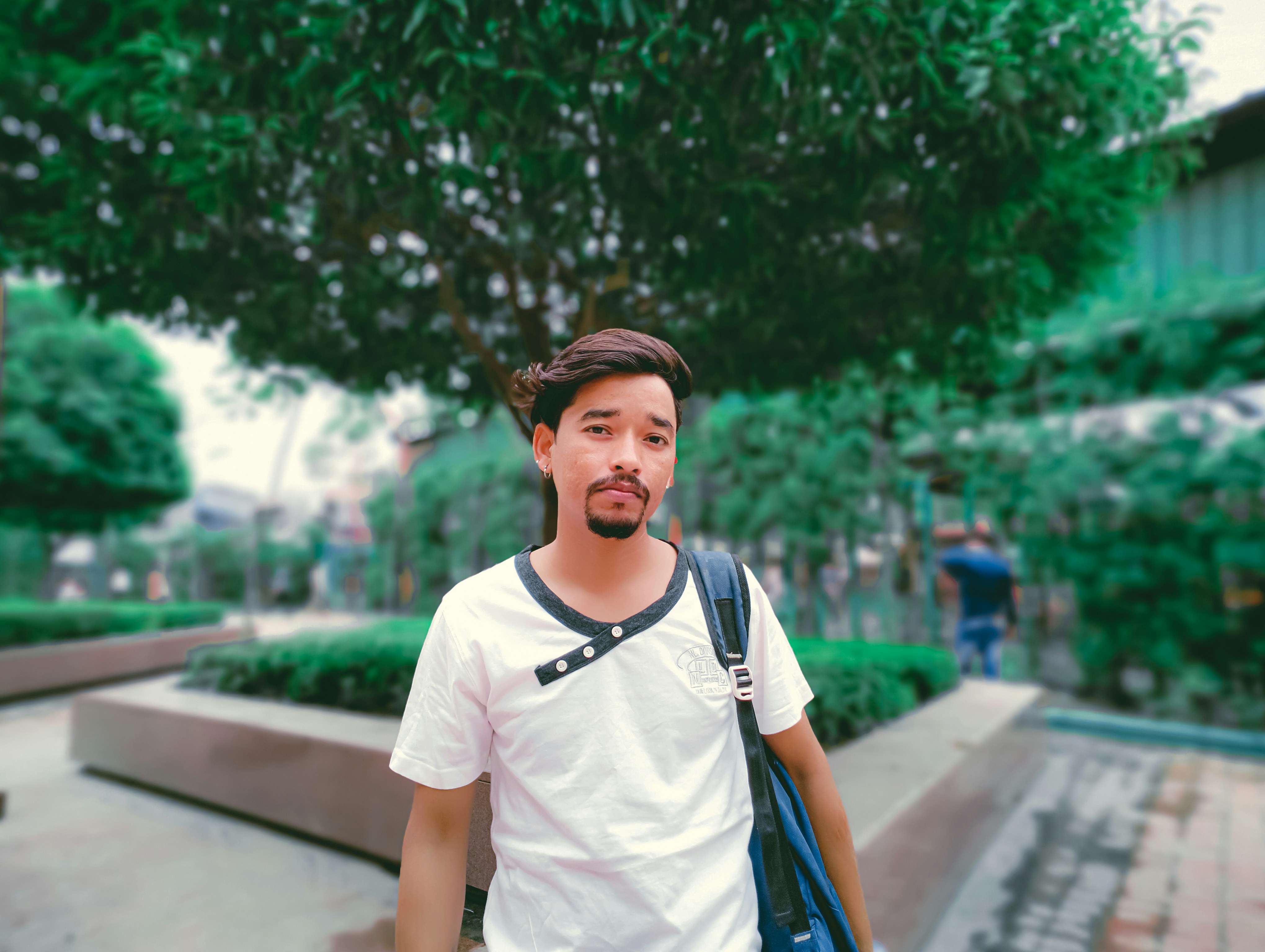 Casually dressed man with a backpack stands in a tree-lined plaza, the subject in sharp focus while the background renders as soft bokeh. This is a street portrait photograph.