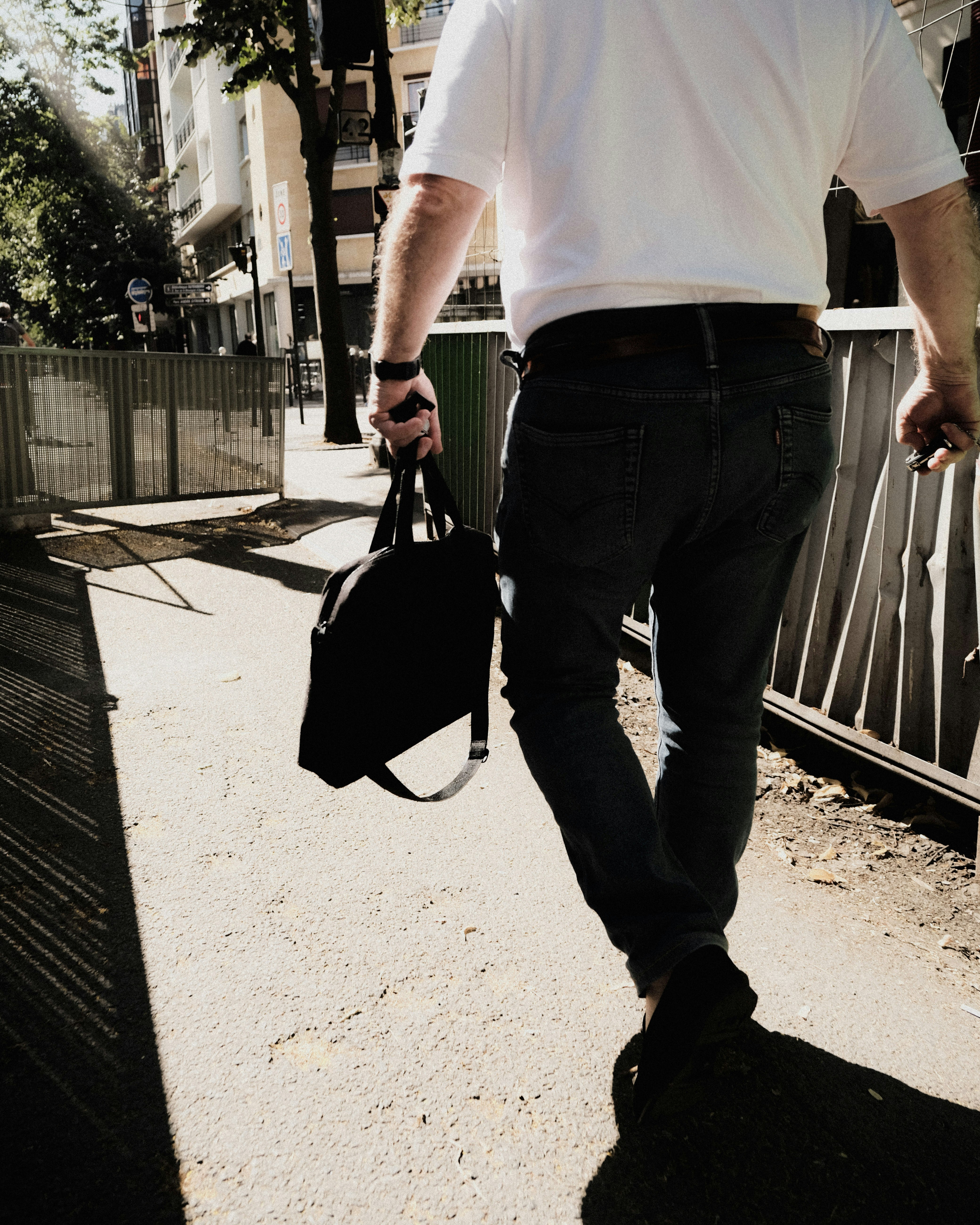 Man walking along a sunlit street, carrying a black bag, with shadows cast by nearby fences and trees.