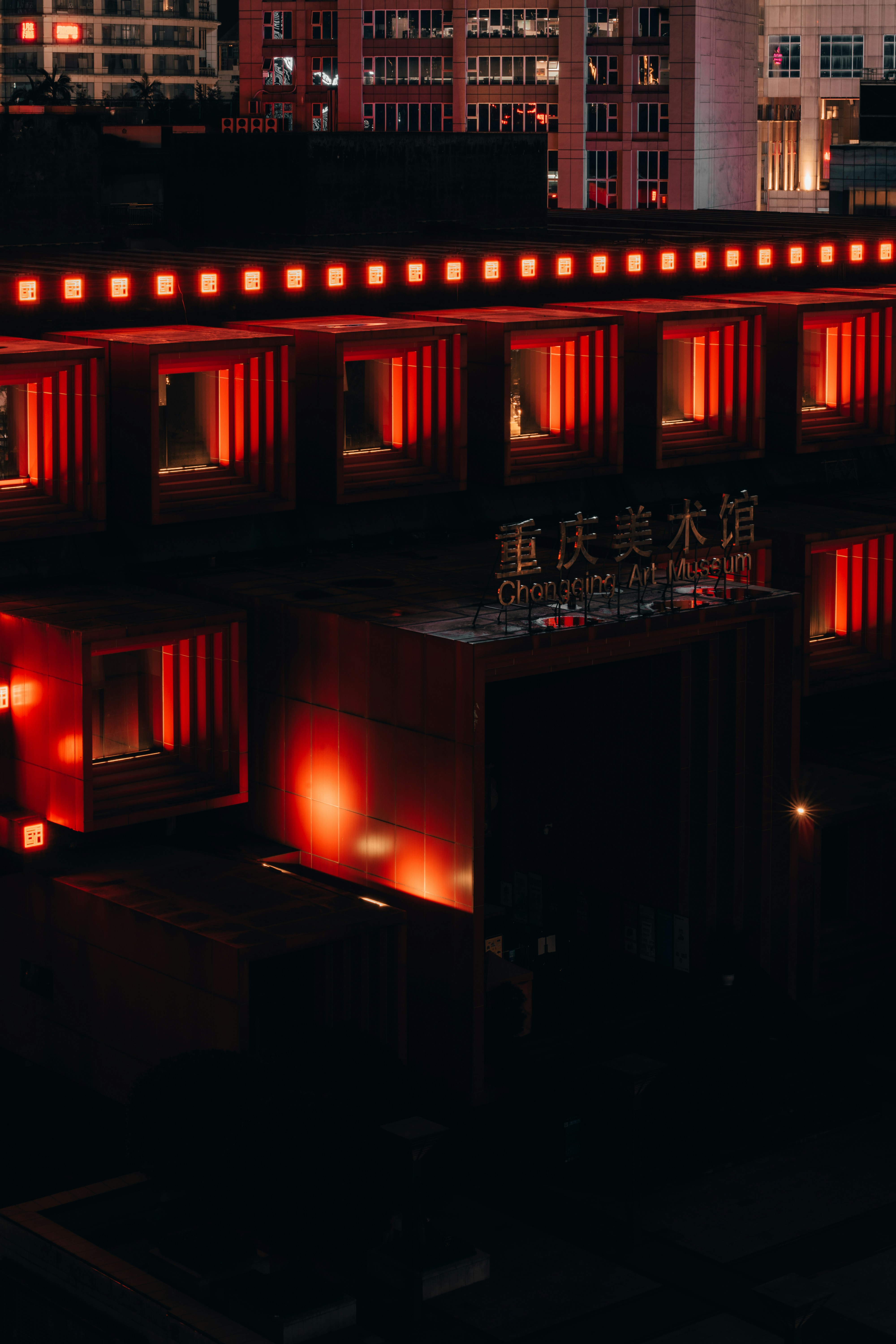 Chongqing Art Museum's striking red exterior illuminated at night, showcasing modern architectural design. The warm glow highlights the unique structure and inviting entrance.
