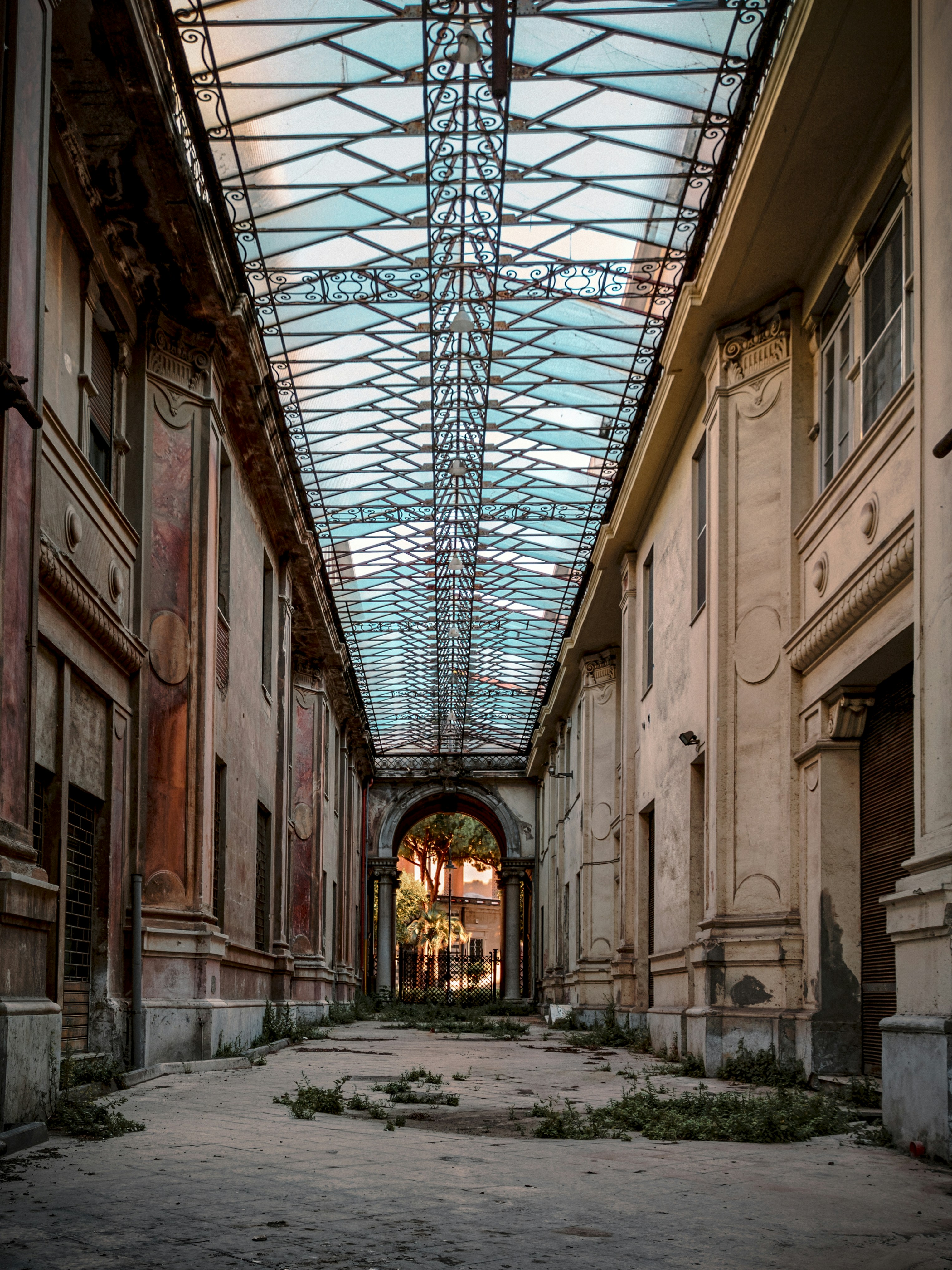 An abandoned corridor with ornate architecture and a glass roof, overgrown with weeds, leading to a distant archway illuminated by soft light.