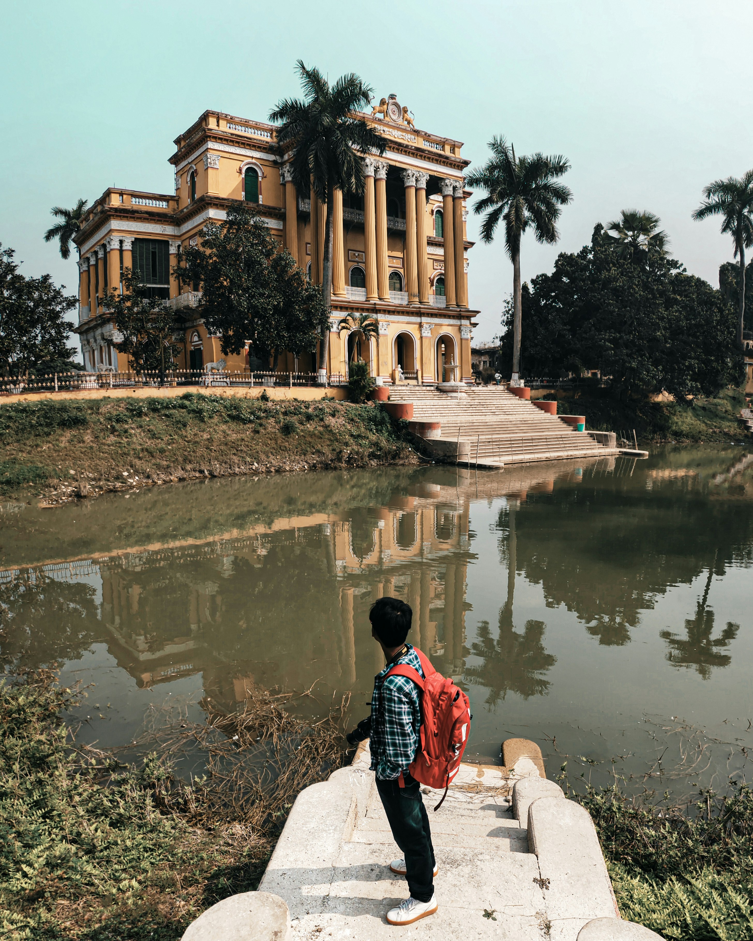 A traveler with a red backpack stands on a stone jetty before a colonial villa reflected in a tranquil pond.