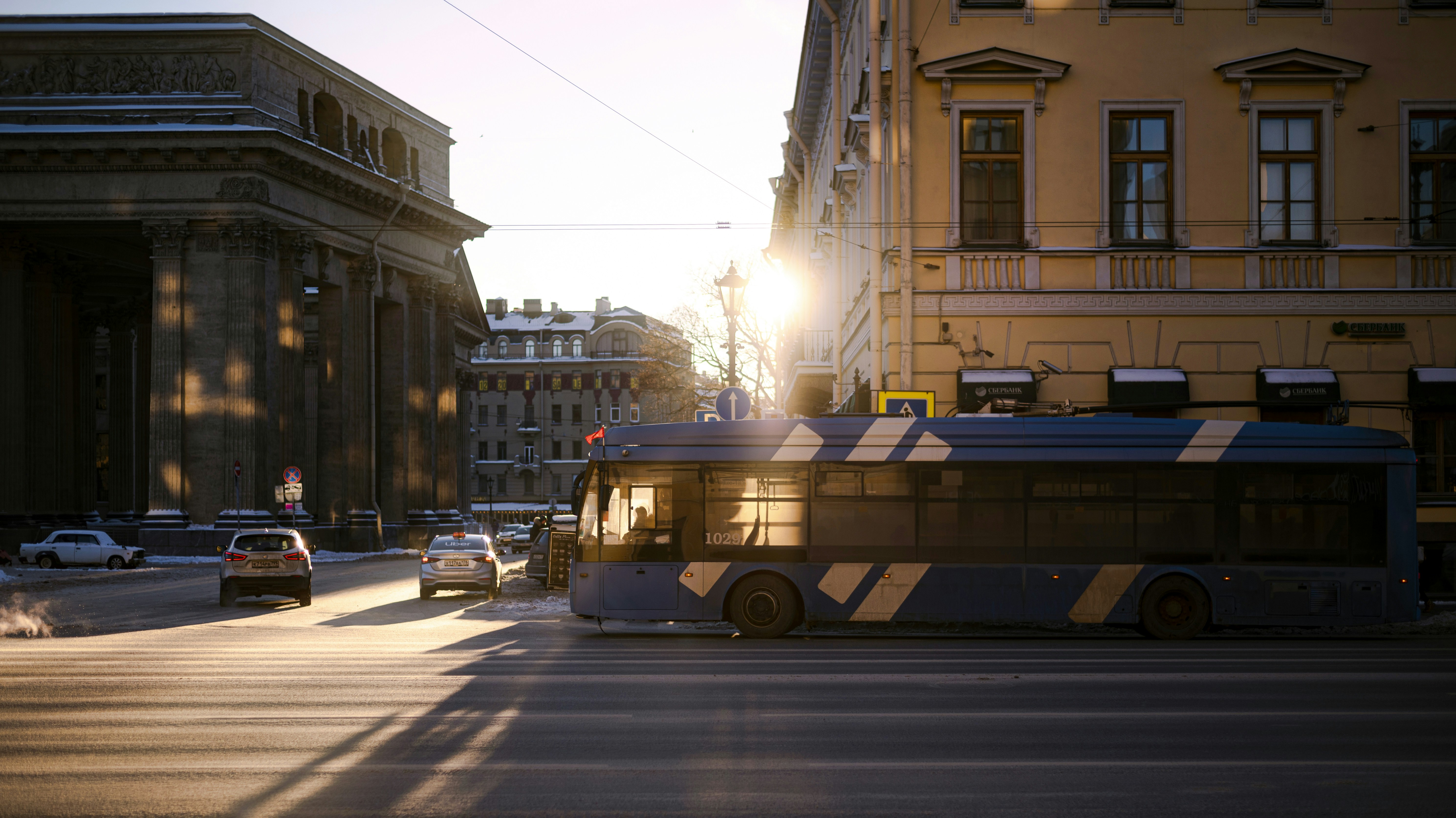 City bus navigating a busy intersection as sunlight filters through buildings, casting long shadows. The scene captures the essence of urban movement.