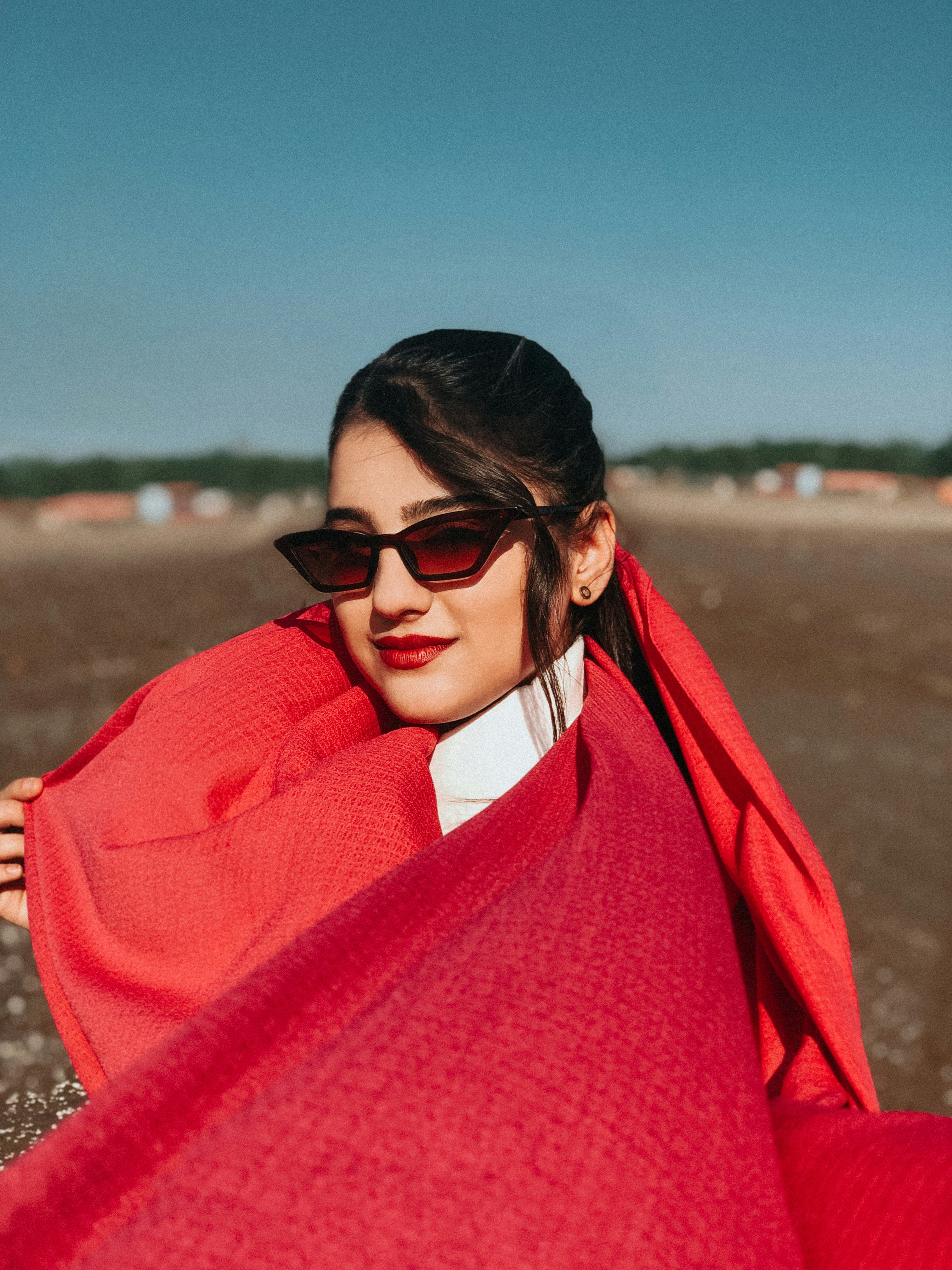 A woman draped in a vivid red shawl, exuding confidence and style against a clear blue sky. Her chic sunglasses complement the bold colors of her attire.