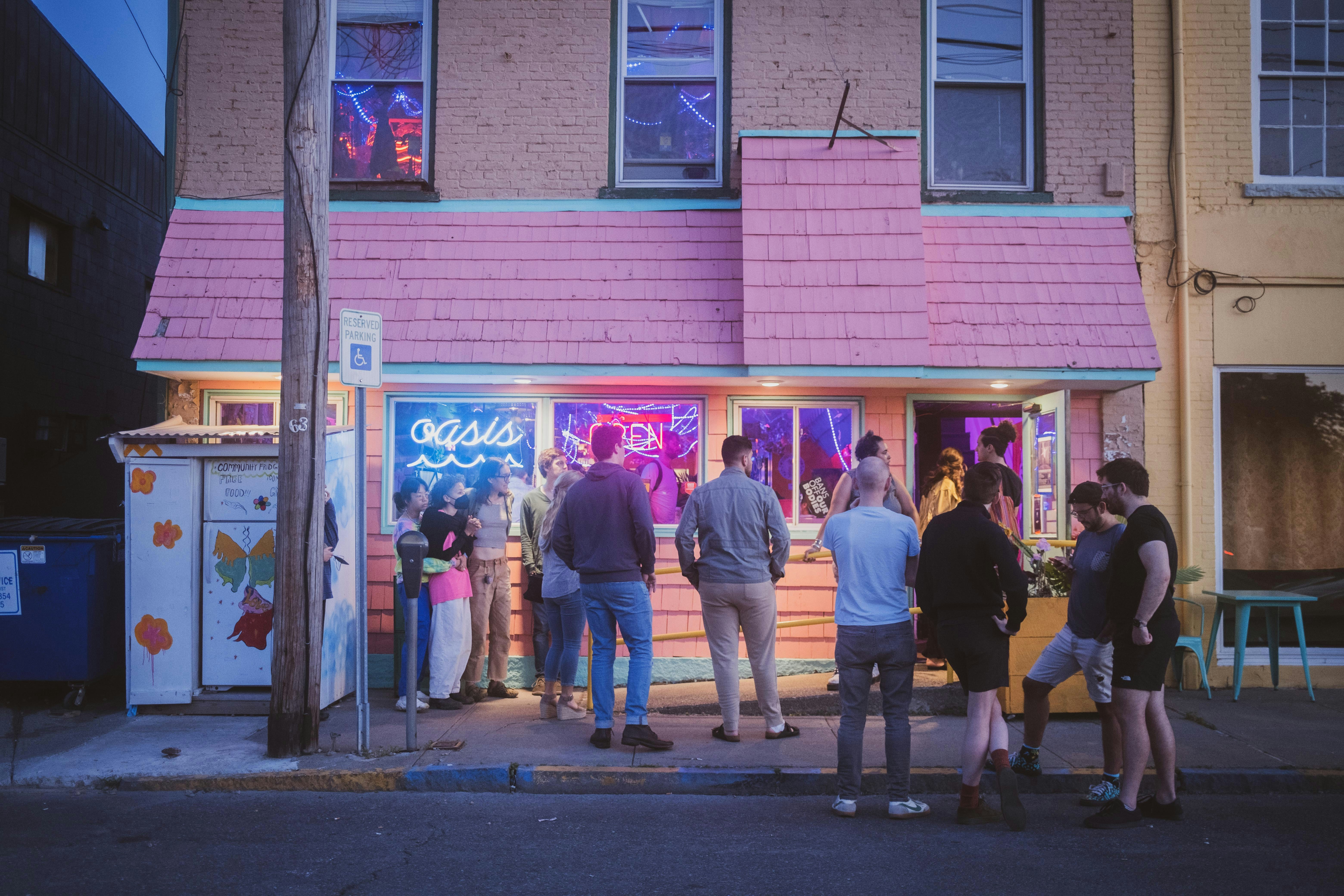 People gather outside a brightly lit street shop under a dusky sky.