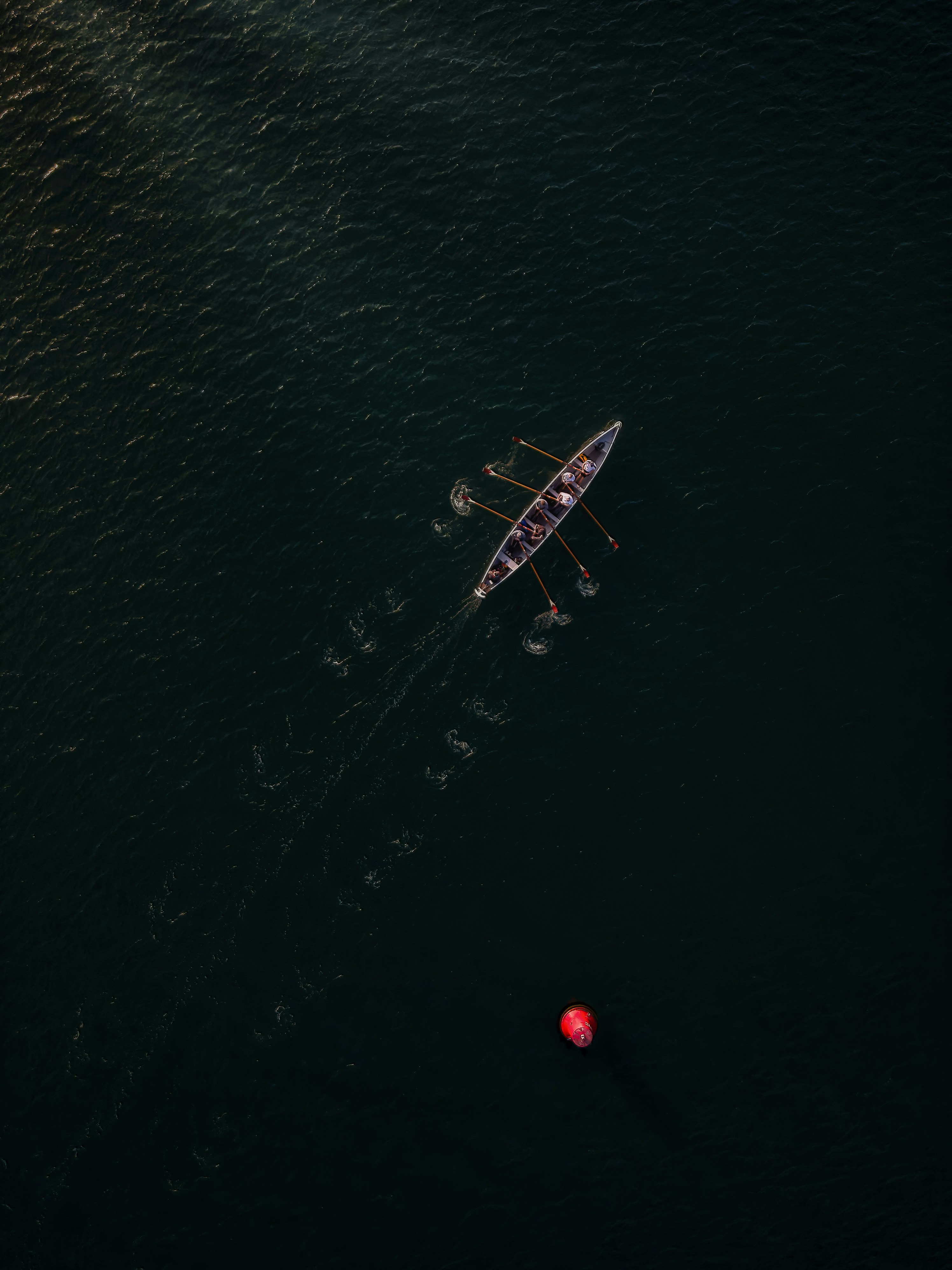 A crew of rowers propels their boat across dark waters, leaving ripples in their wake, with a bright buoy nearby.