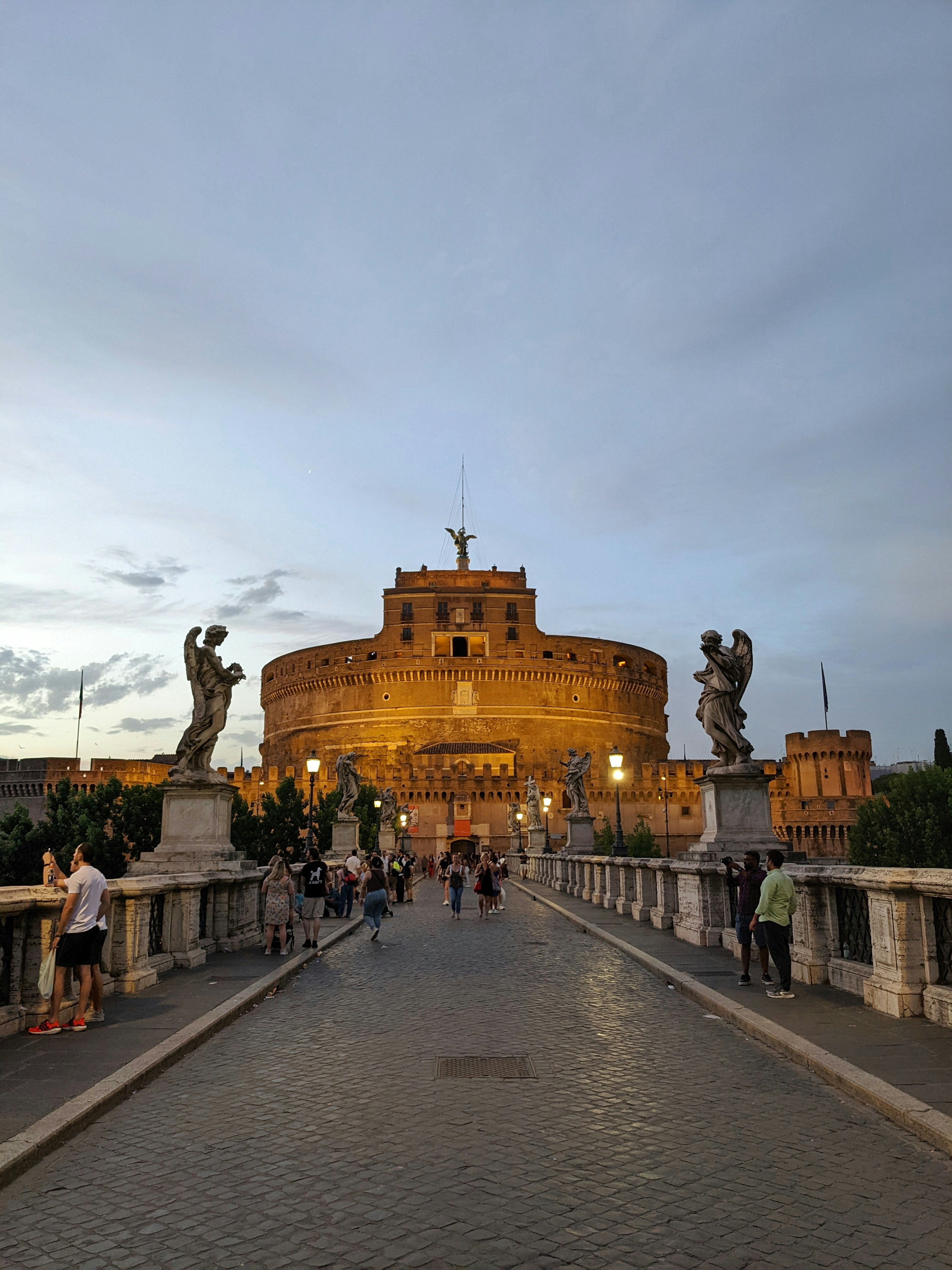 Historic Castel Sant'Angelo illuminated at dusk, seen from the bridge adorned with statues and bustling with visitors.