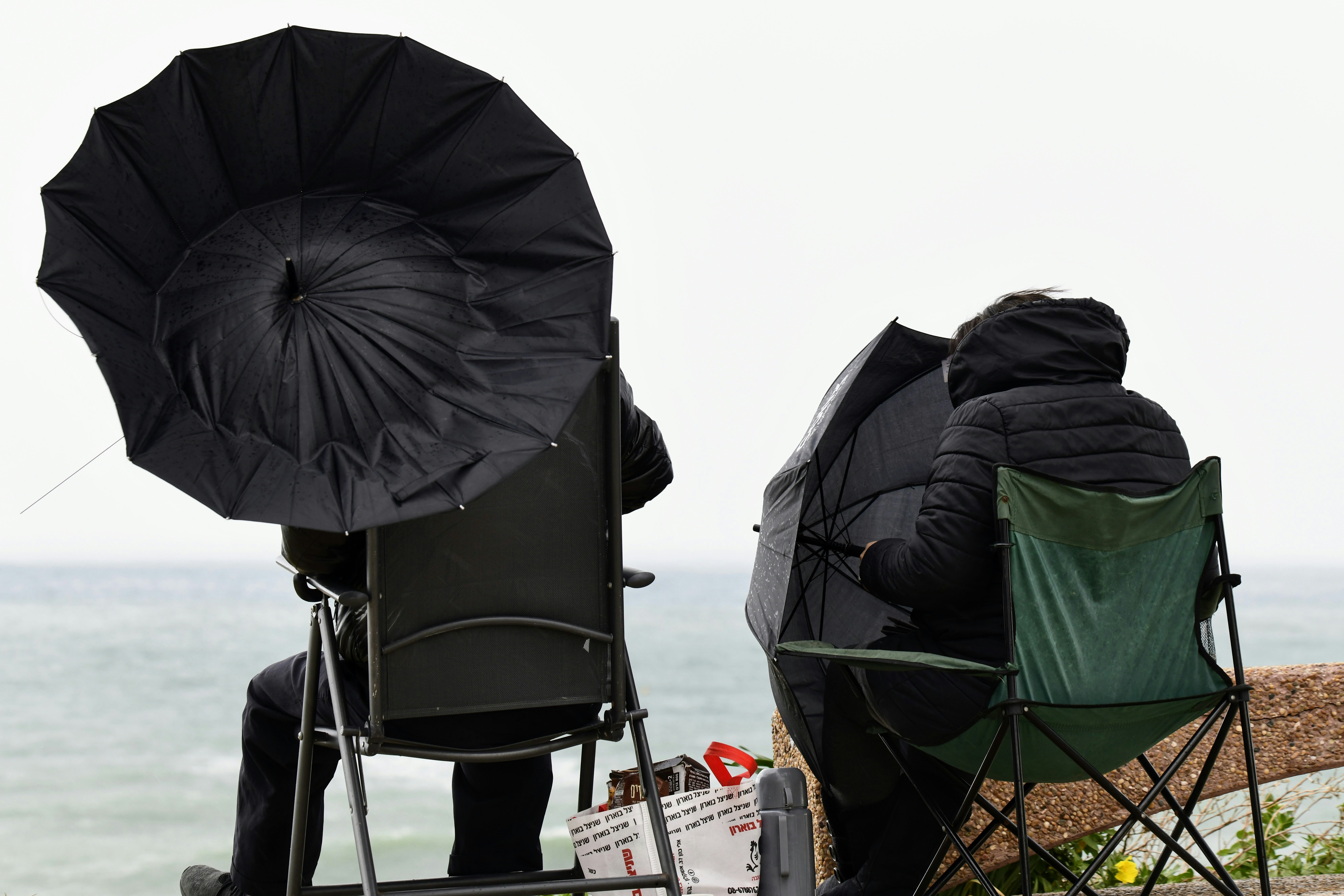 Two figures seated in folding chairs, sheltered by black umbrellas, gaze toward the tranquil sea. The scene captures a moment of quiet reflection by the water's edge.