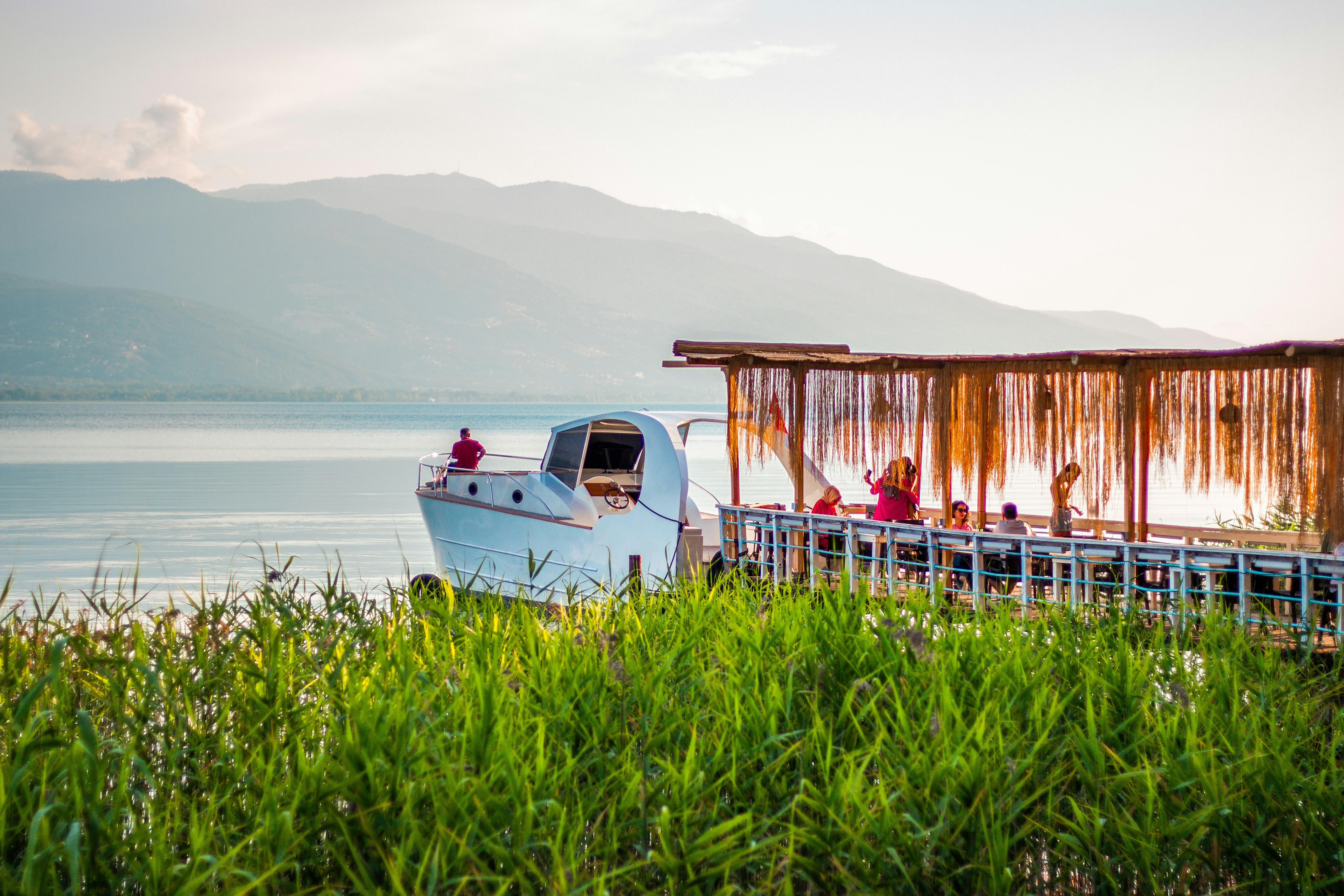 A leisure boat docked at a wooden pier surrounded by lush greenery, with people enjoying the tranquil lakeside atmosphere. The mountains loom softly in the background.