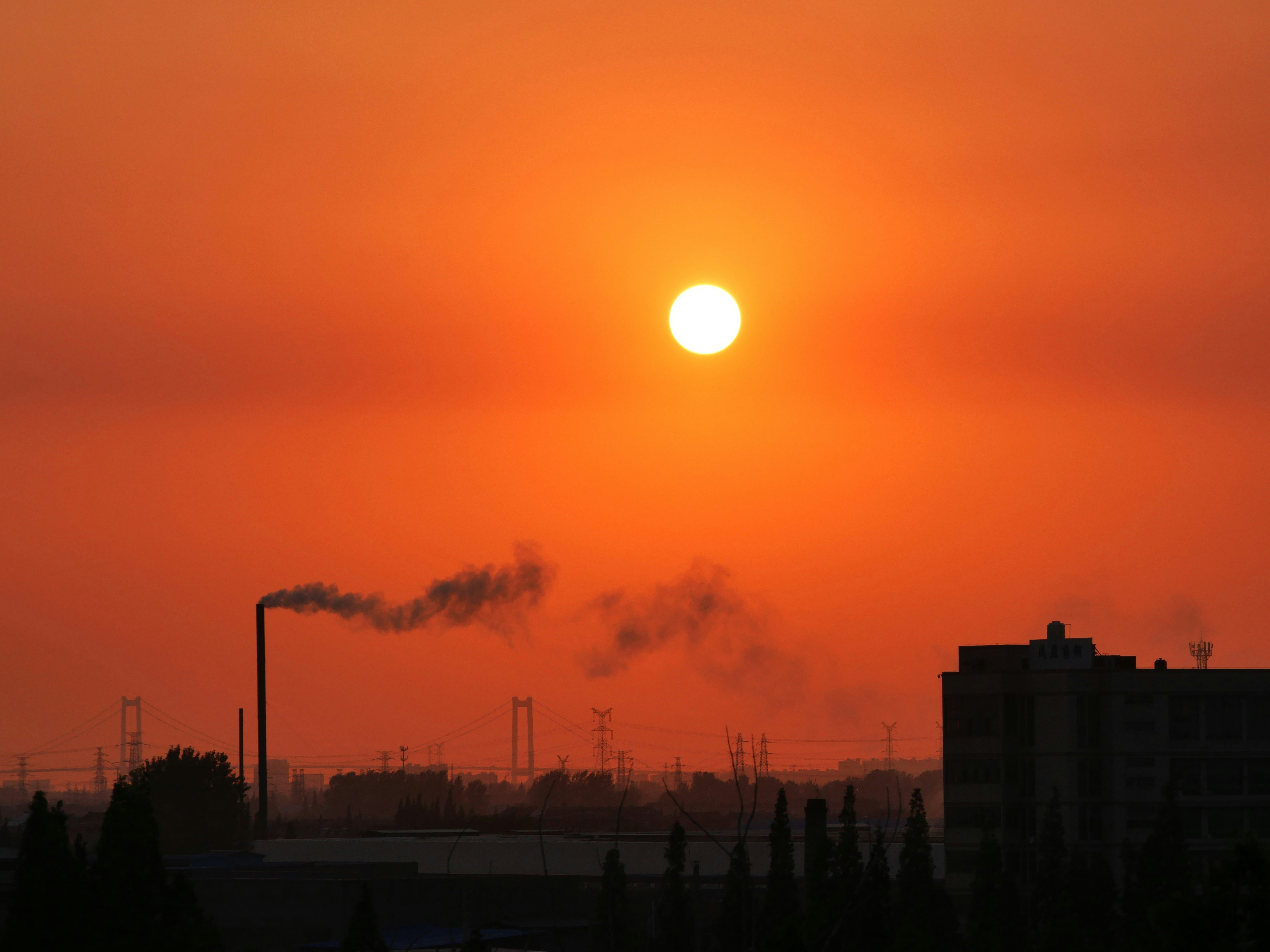 Silhouetted industrial landscape against a vibrant sunset, featuring a smokestack emitting wisps of smoke. The scene captures the contrast between nature and industrialization.