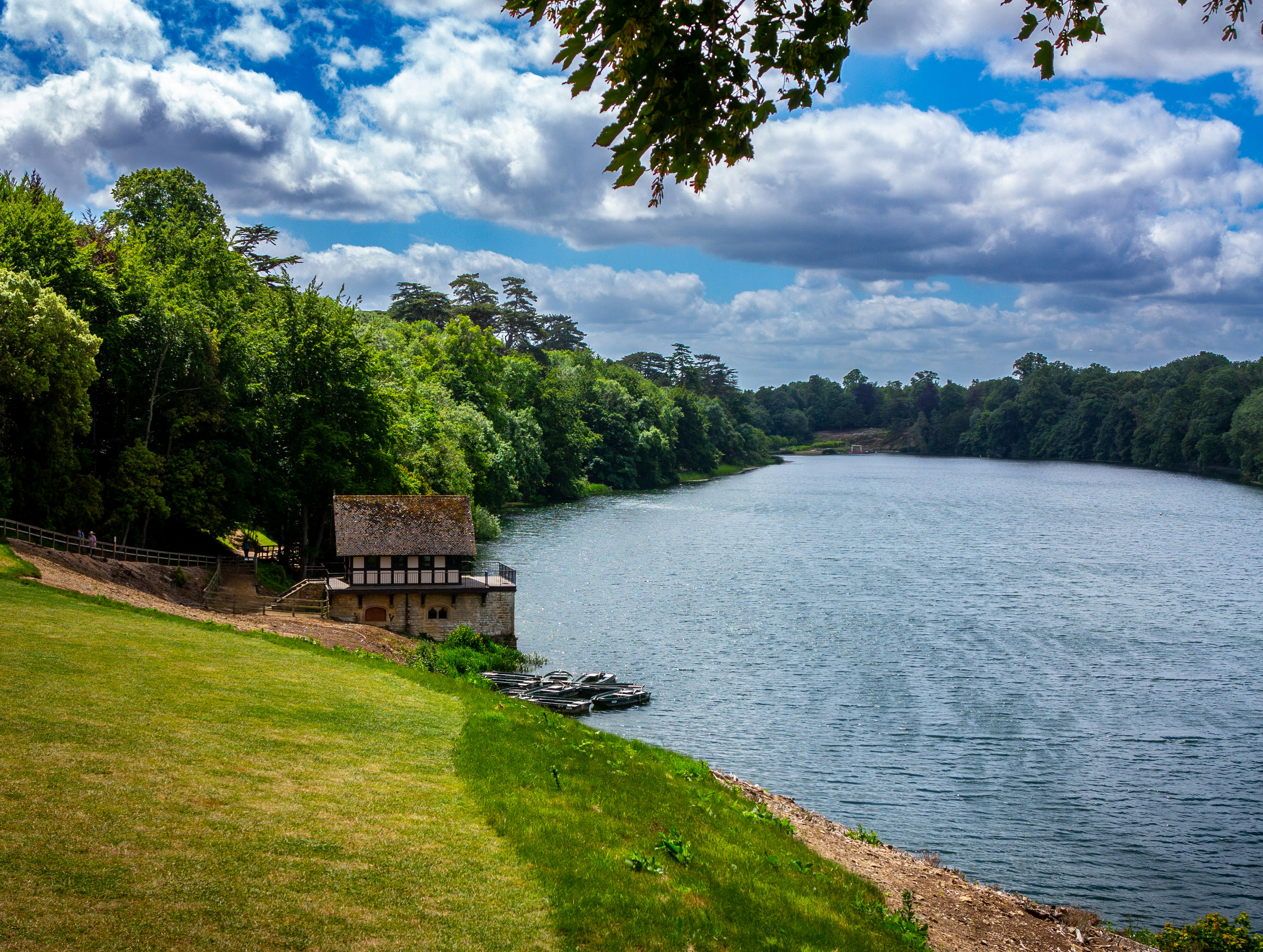 Cottage nestled on a grassy hill beside a tranquil river, surrounded by lush trees and a partly cloudy sky.