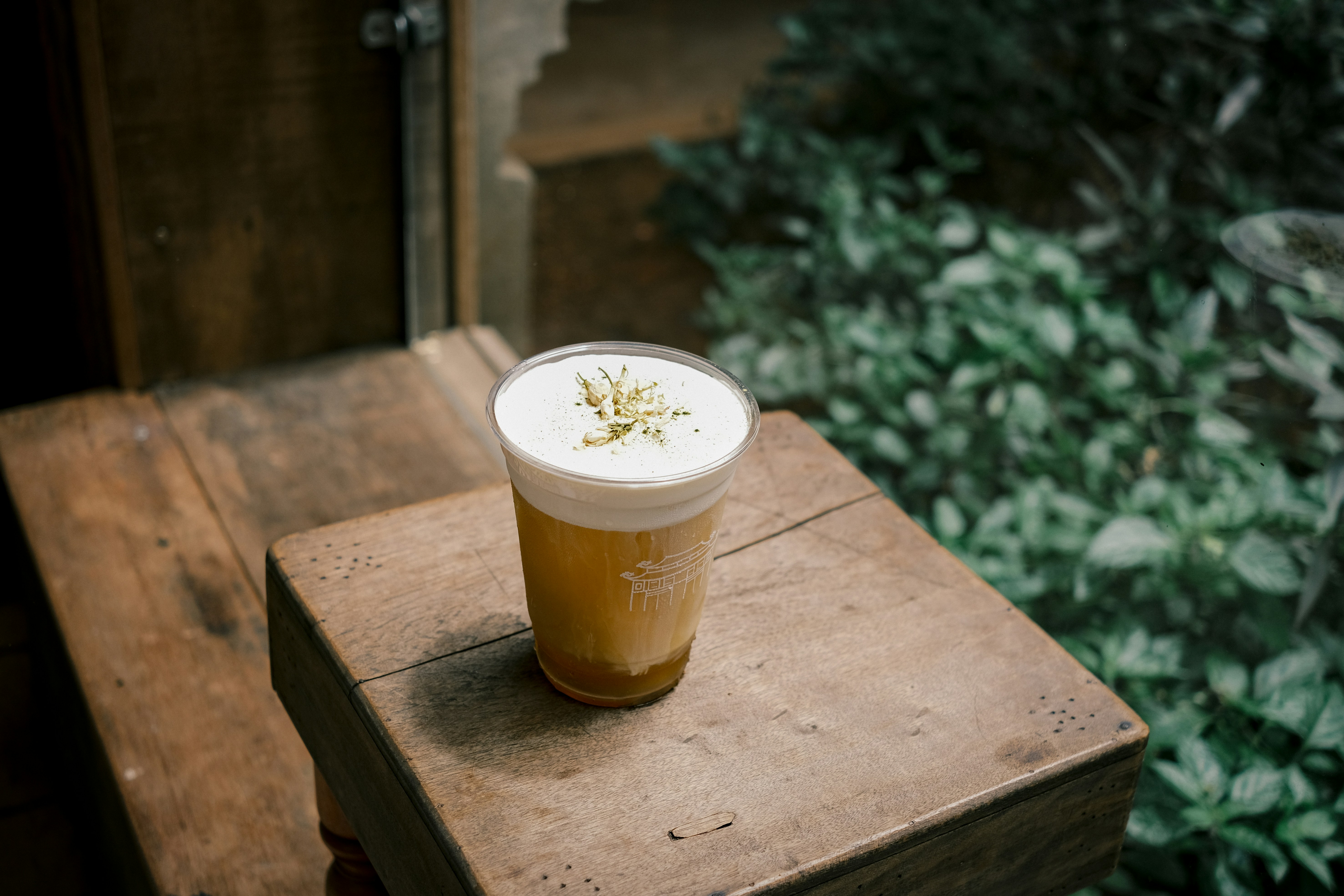 A frothy beverage garnished with herbs rests on a rustic wooden table, surrounded by lush greenery. The setting exudes a warm, inviting atmosphere.