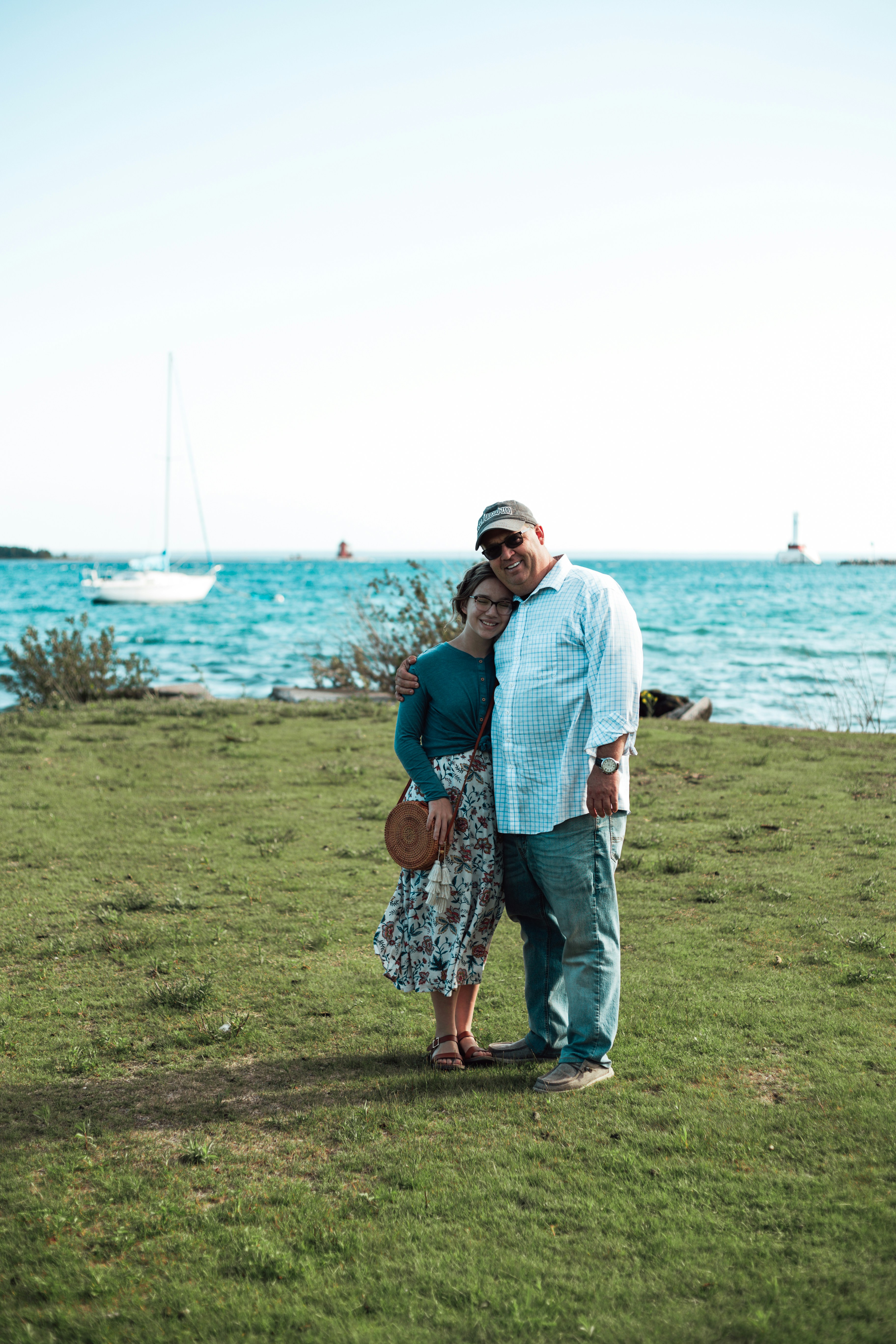 Couple sharing a joyful moment on a grassy shoreline with a calm sea and sailboats in the background.