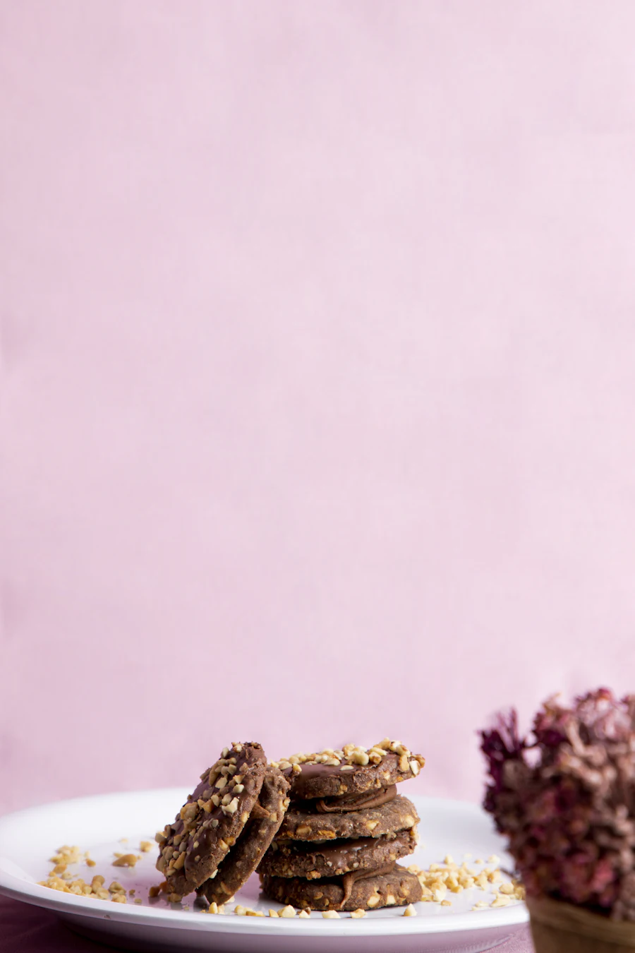Stack of chocolate cookies sprinkled with chopped nuts on a white plate against a soft pink background, with a blurred plant at the right edge.
