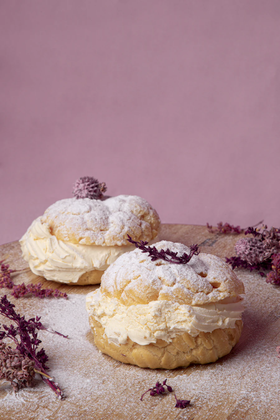 Two cream-filled pastries dusted with powdered sugar on a wooden surface, accented by small purple flowers against a pink backdrop.