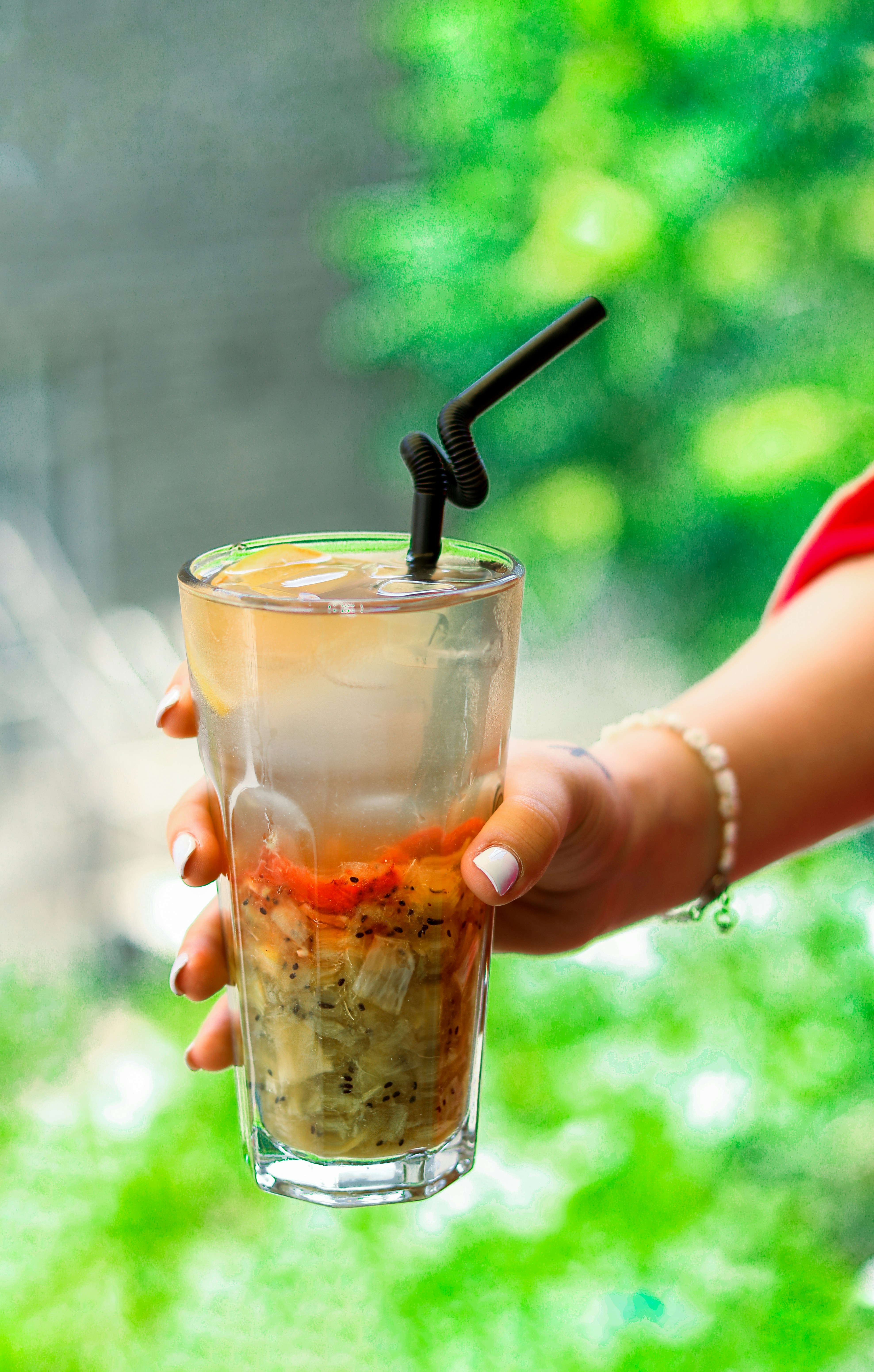 A hand holding a transparent glass filled with a colorful layered drink, featuring fruit garnishes and a black straw against a blurred green background.