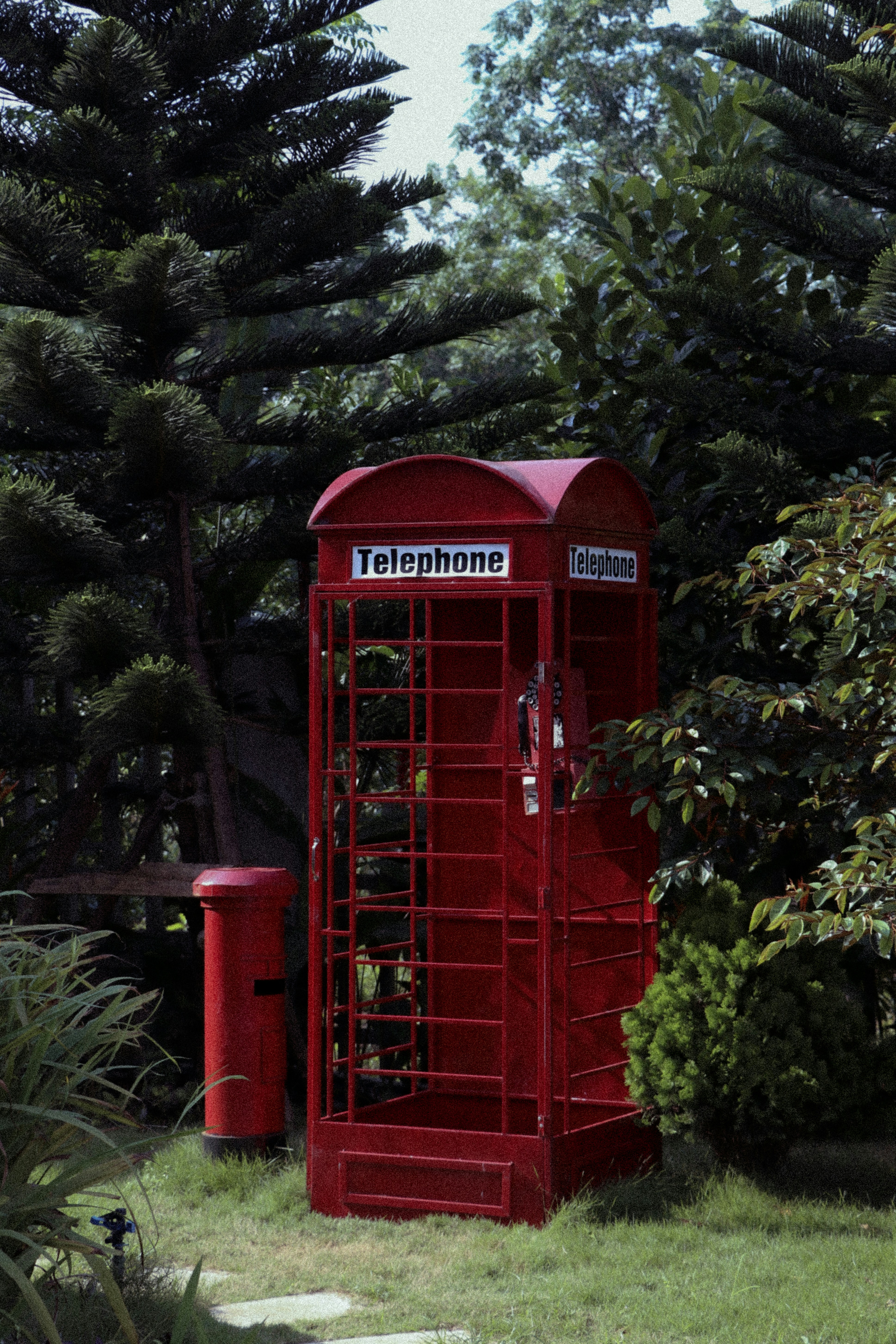 A vintage red telephone booth stands amidst lush greenery, accompanied by a matching postbox, evoking nostalgia and a sense of history.