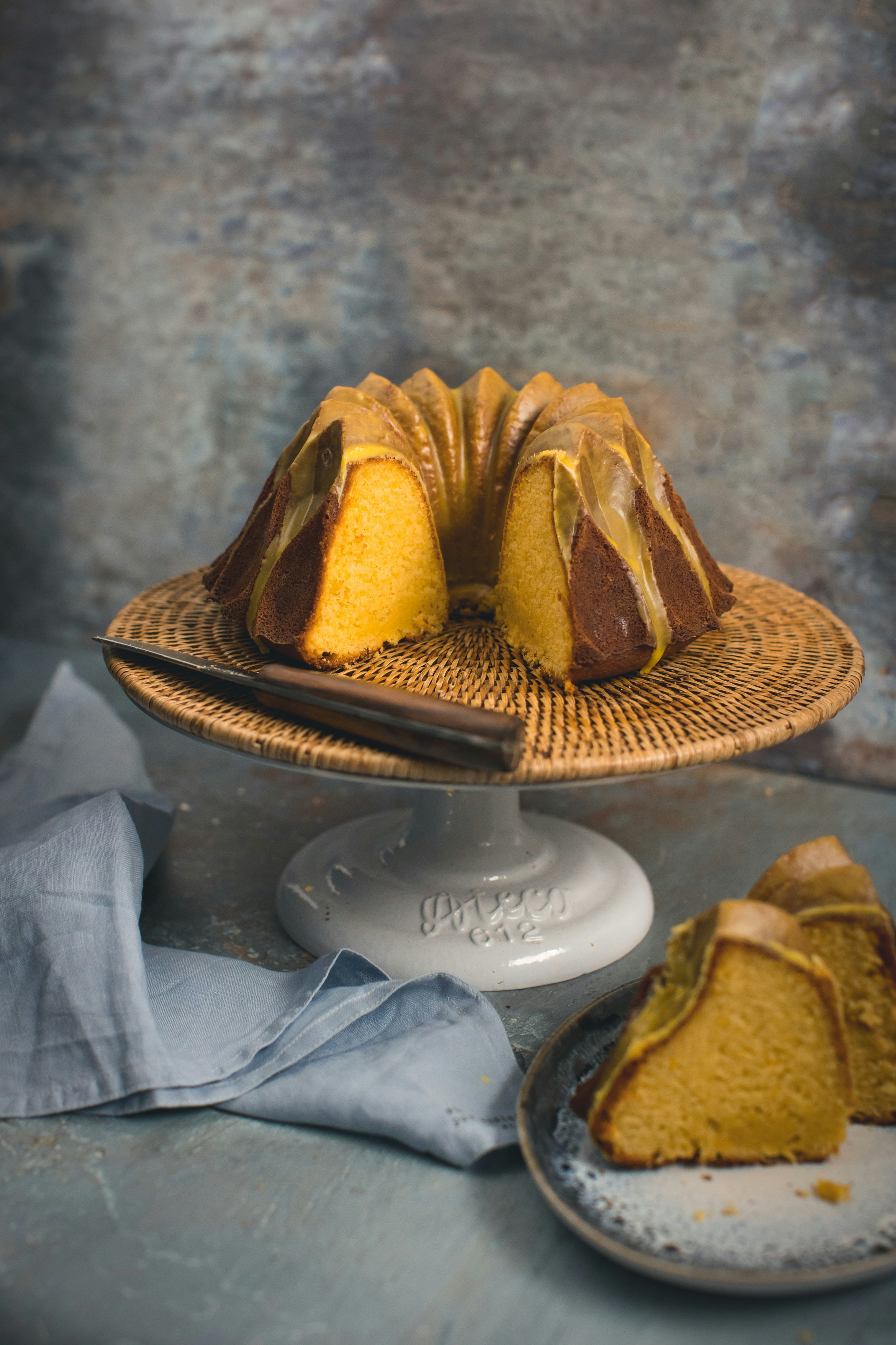 Bundt cake elegantly displayed on a woven platter, accompanied by a slice on a plate. Soft blue linen adds a touch of warmth.