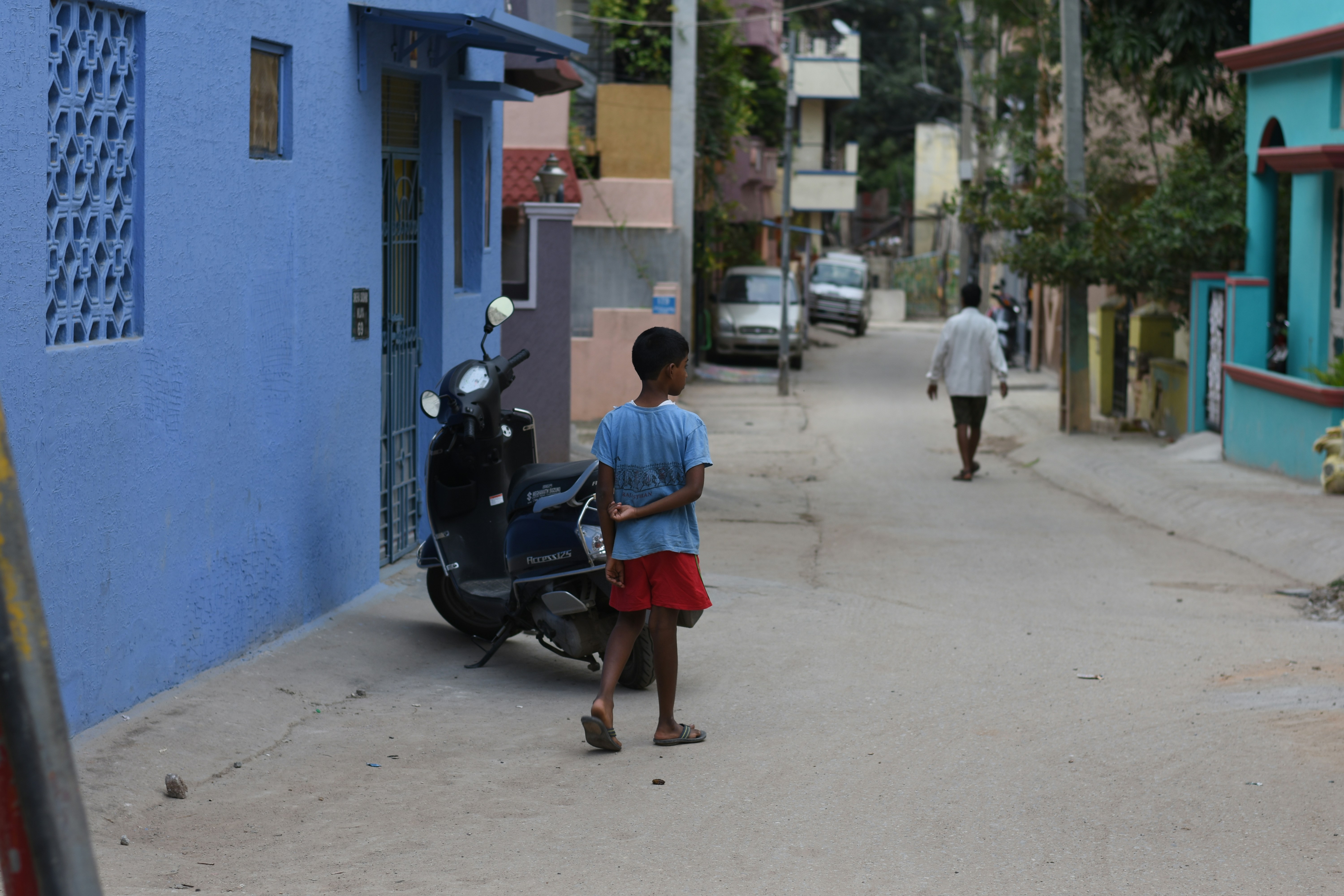 Child in blue shirt and red shorts leads a quiet street toward a distant passerby