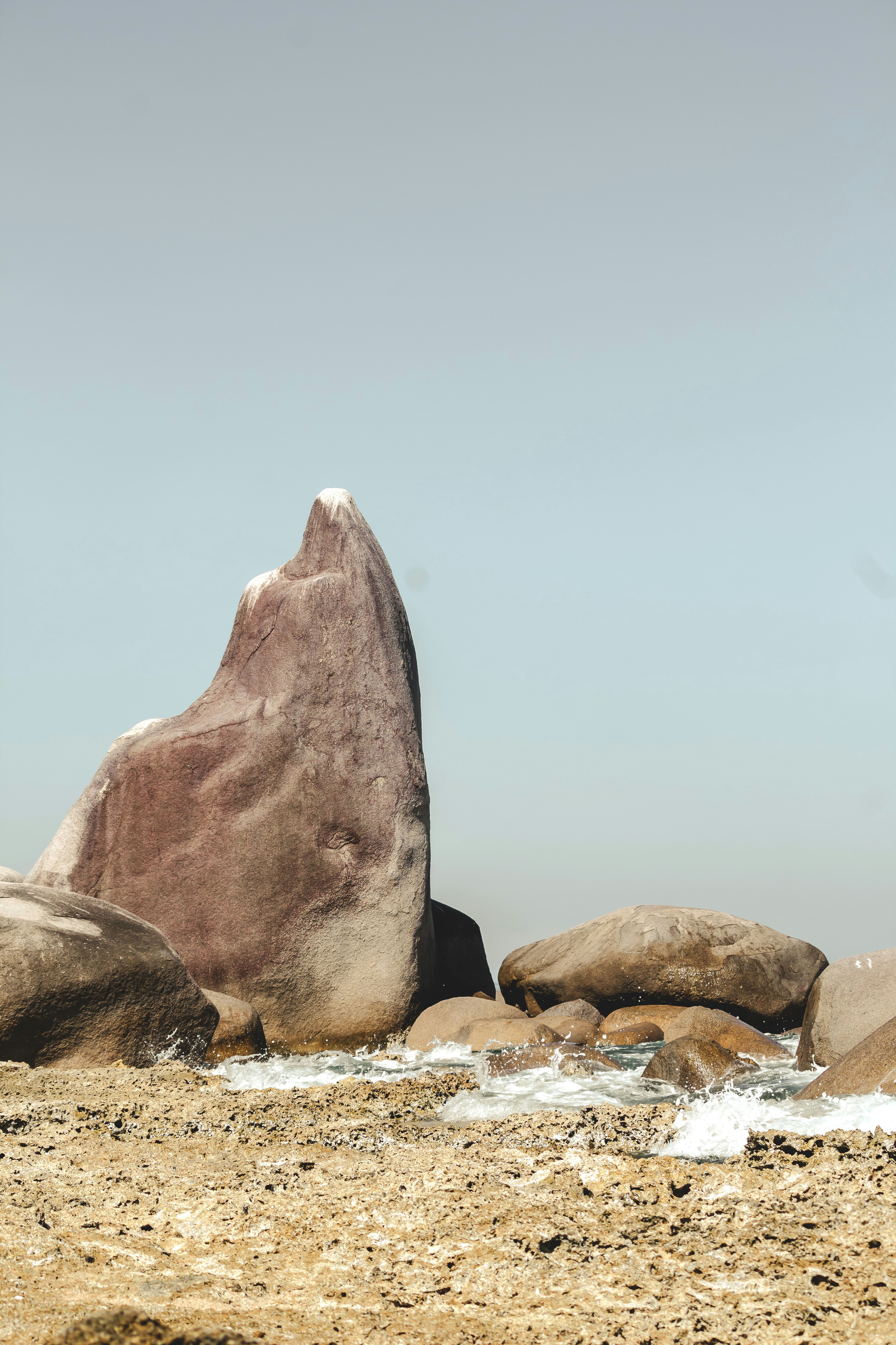 A towering rock formation stands majestically against a clear sky, surrounded by smaller stones and gentle waves lapping at the shore.