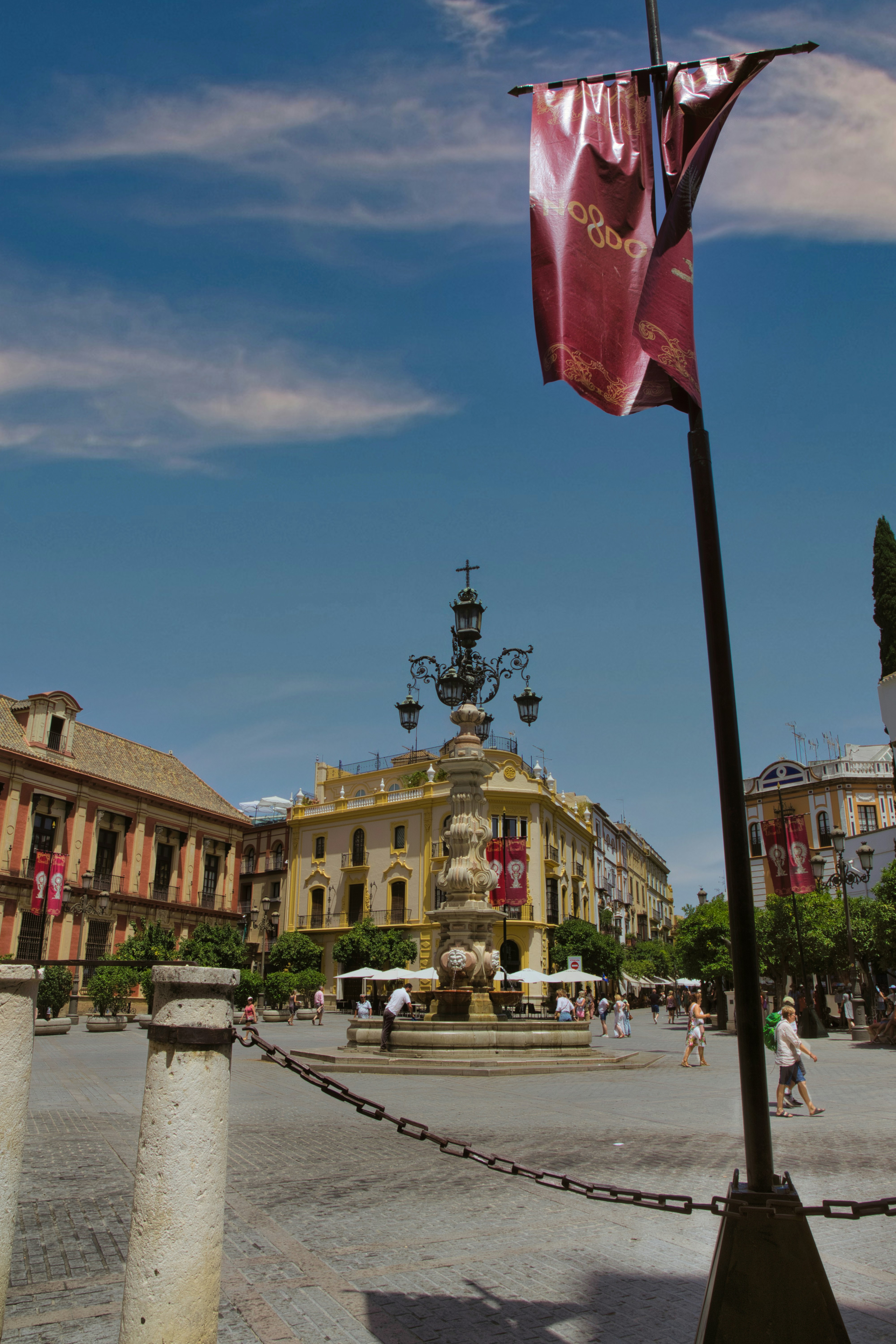 Charming plaza featuring a central fountain surrounded by vibrant architecture and lively pedestrians. Banners flutter in the breeze, adding to the scene's dynamic atmosphere.