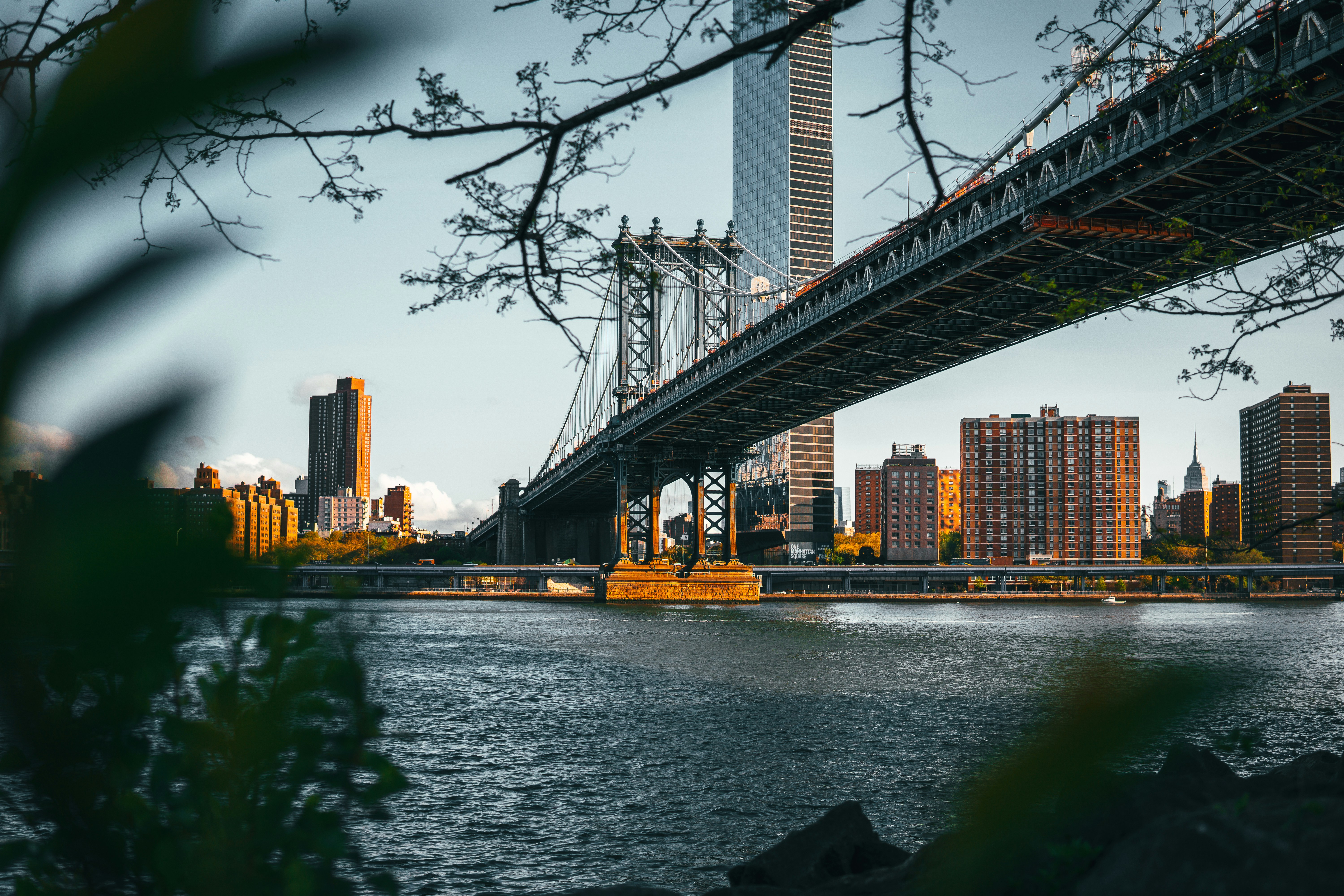 Brooklyn Bridge arching over the East River with city skyline in the background, framed by lush greenery.