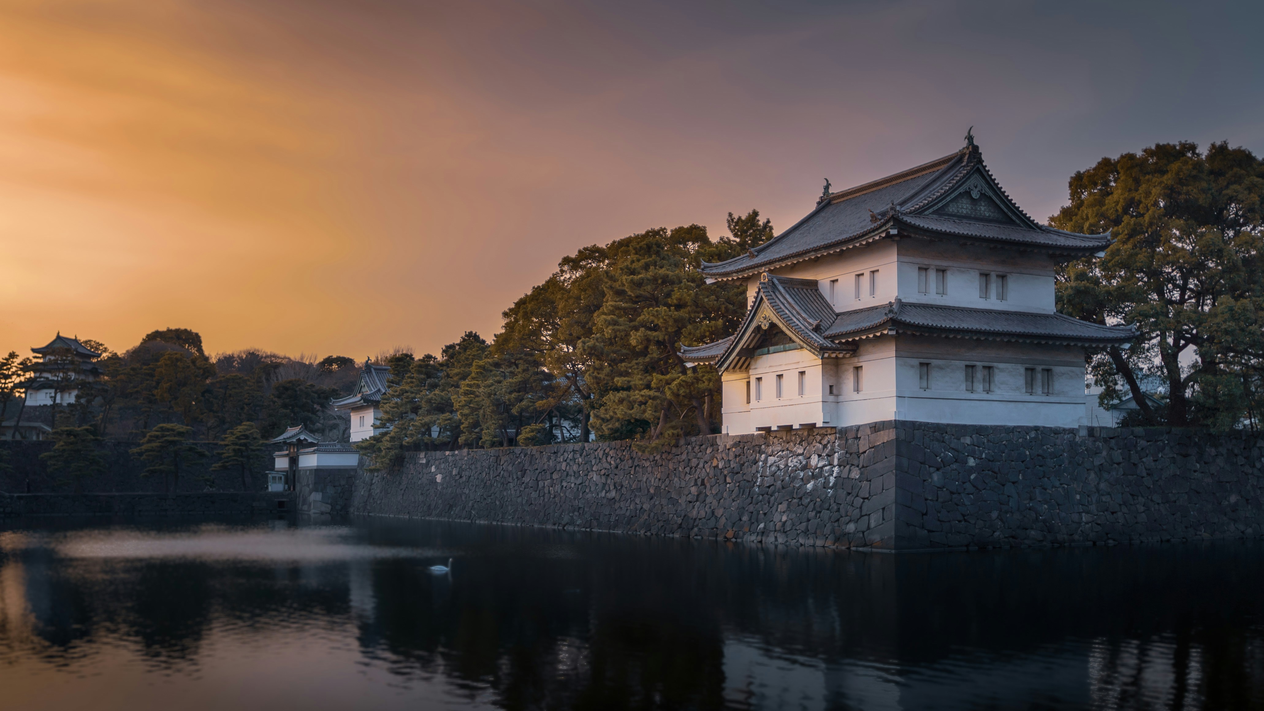 Traditional Japanese castle reflecting in still waters during sunset, surrounded by lush greenery.