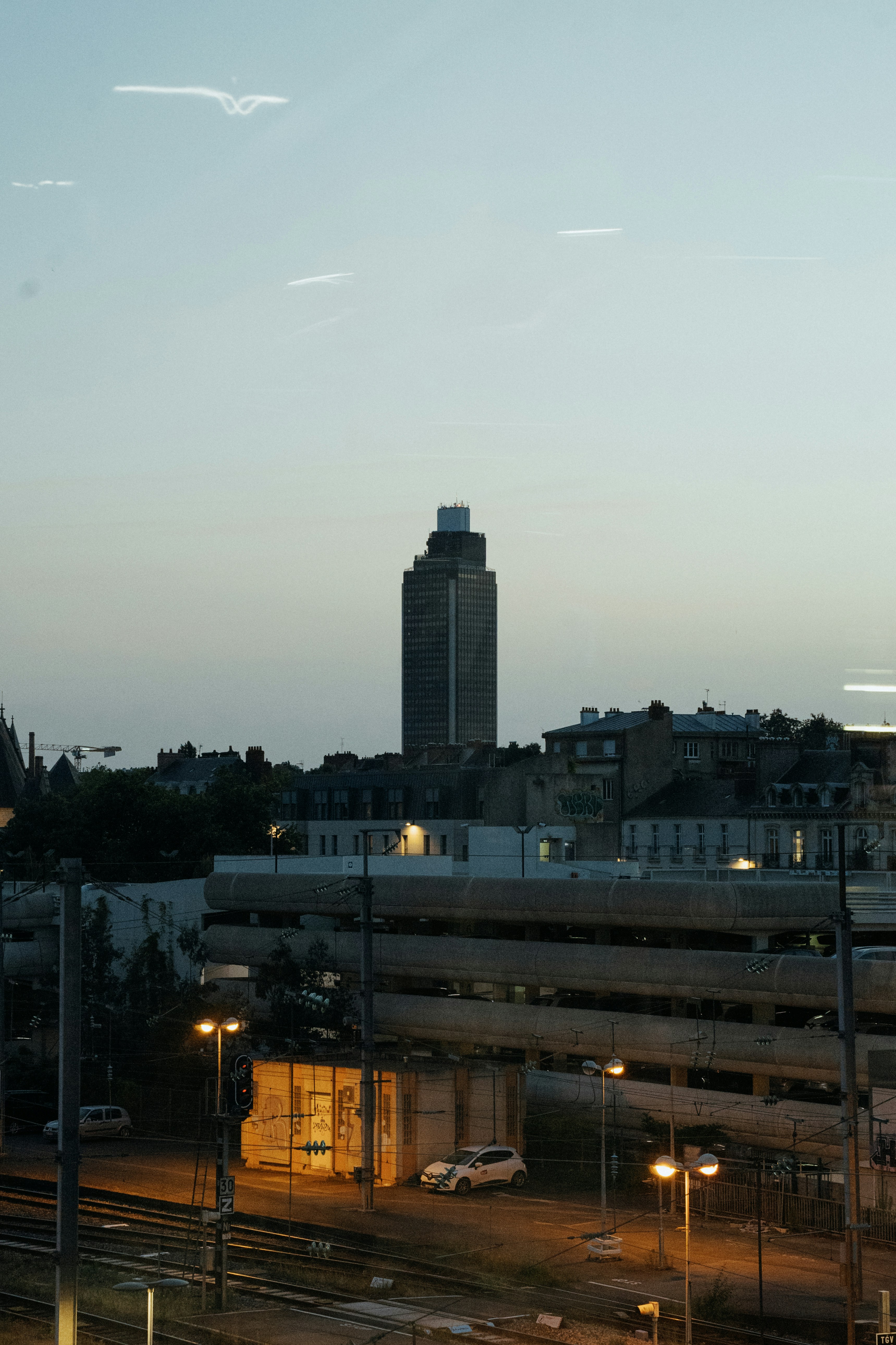 Towering skyscraper dominates the skyline as twilight descends over the cityscape, framed by rooftops and rail lines.