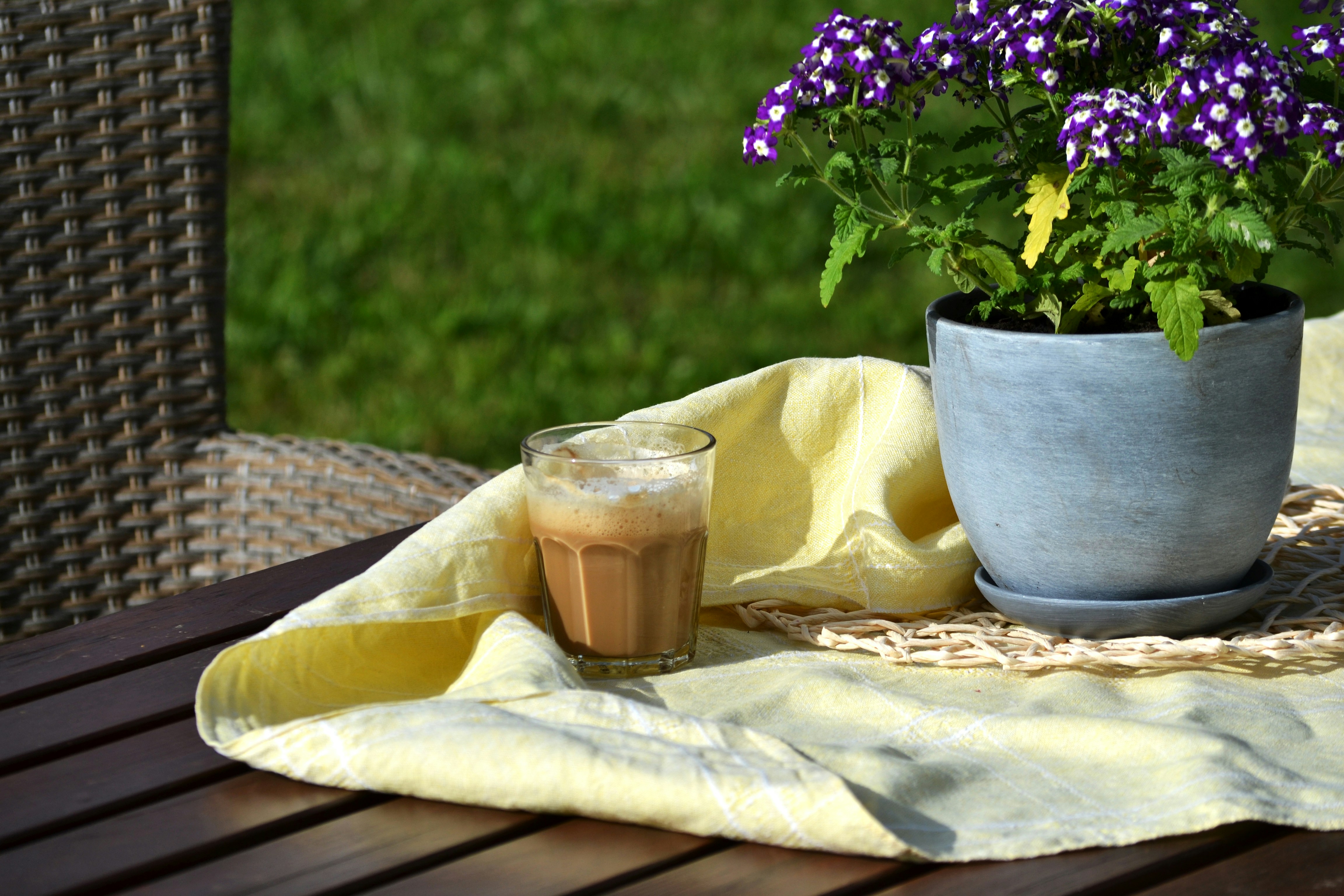A glass of iced coffee sits on a sunlit wooden table, accompanied by a vibrant flower pot and a soft yellow cloth.