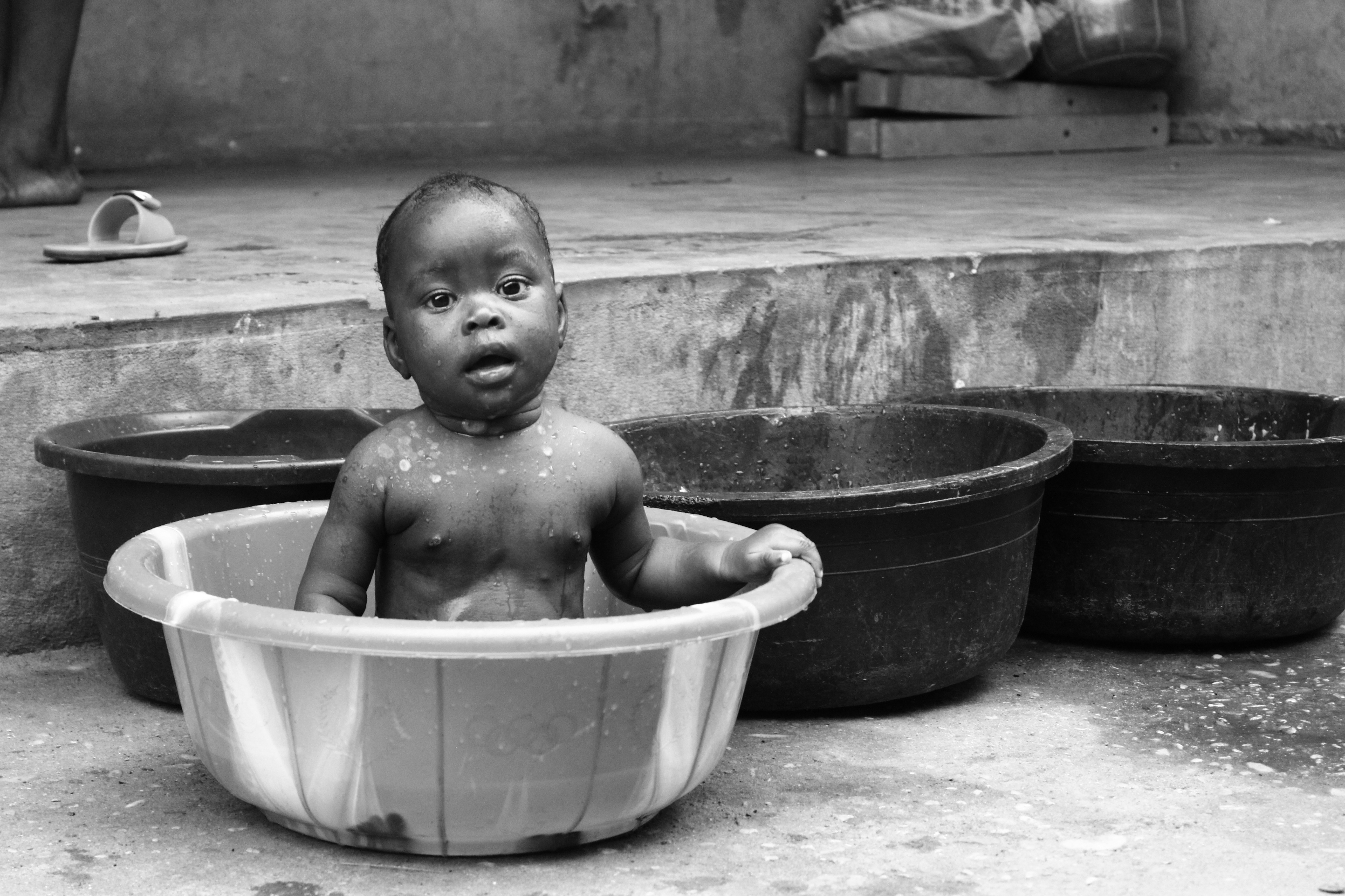 Baby sitting in a small tub with water droplets on skin, surrounded by larger basins on a concrete floor.