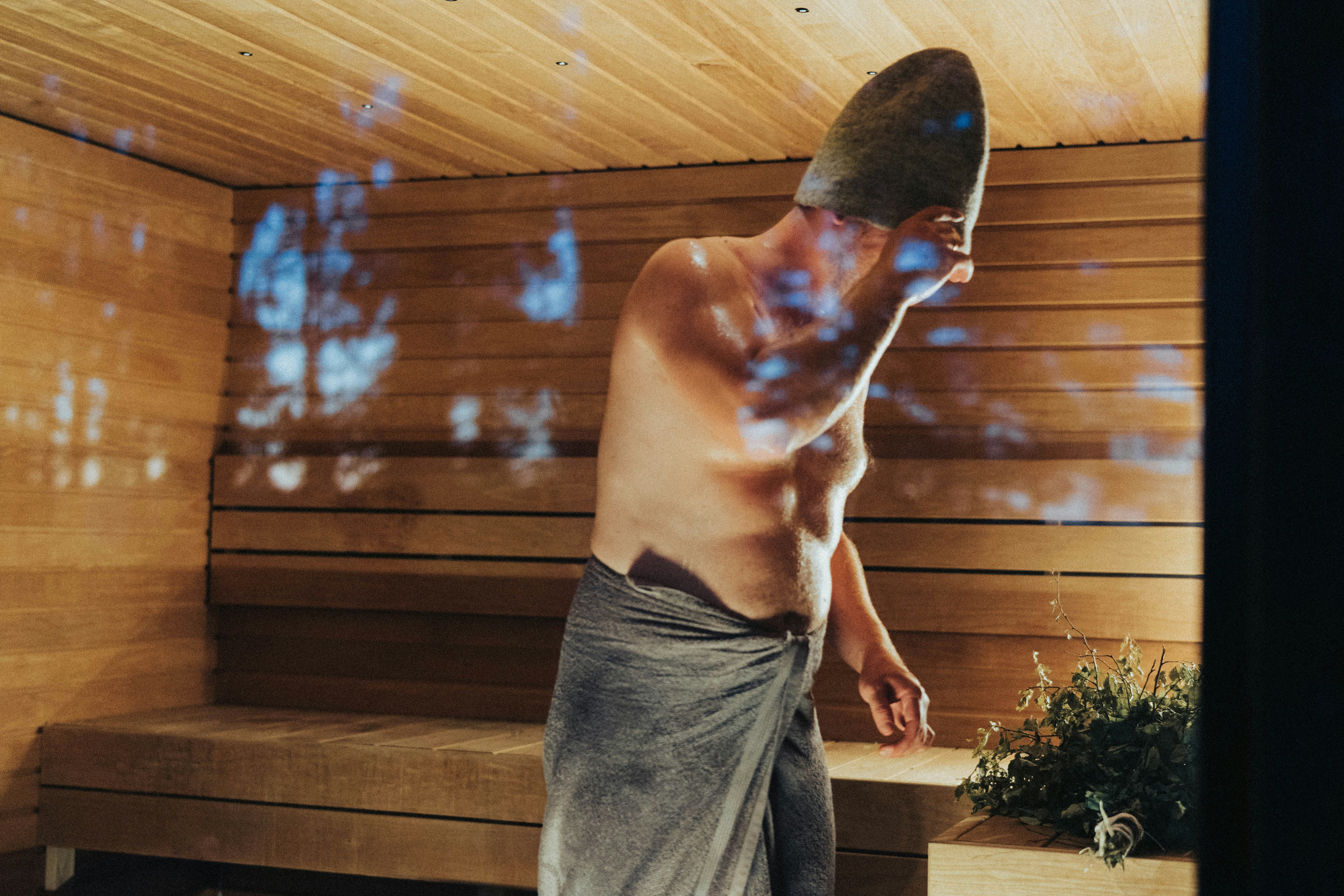 A man in a sauna, partially covered with a towel, interacts with the serene environment, surrounded by wooden benches and greenery.