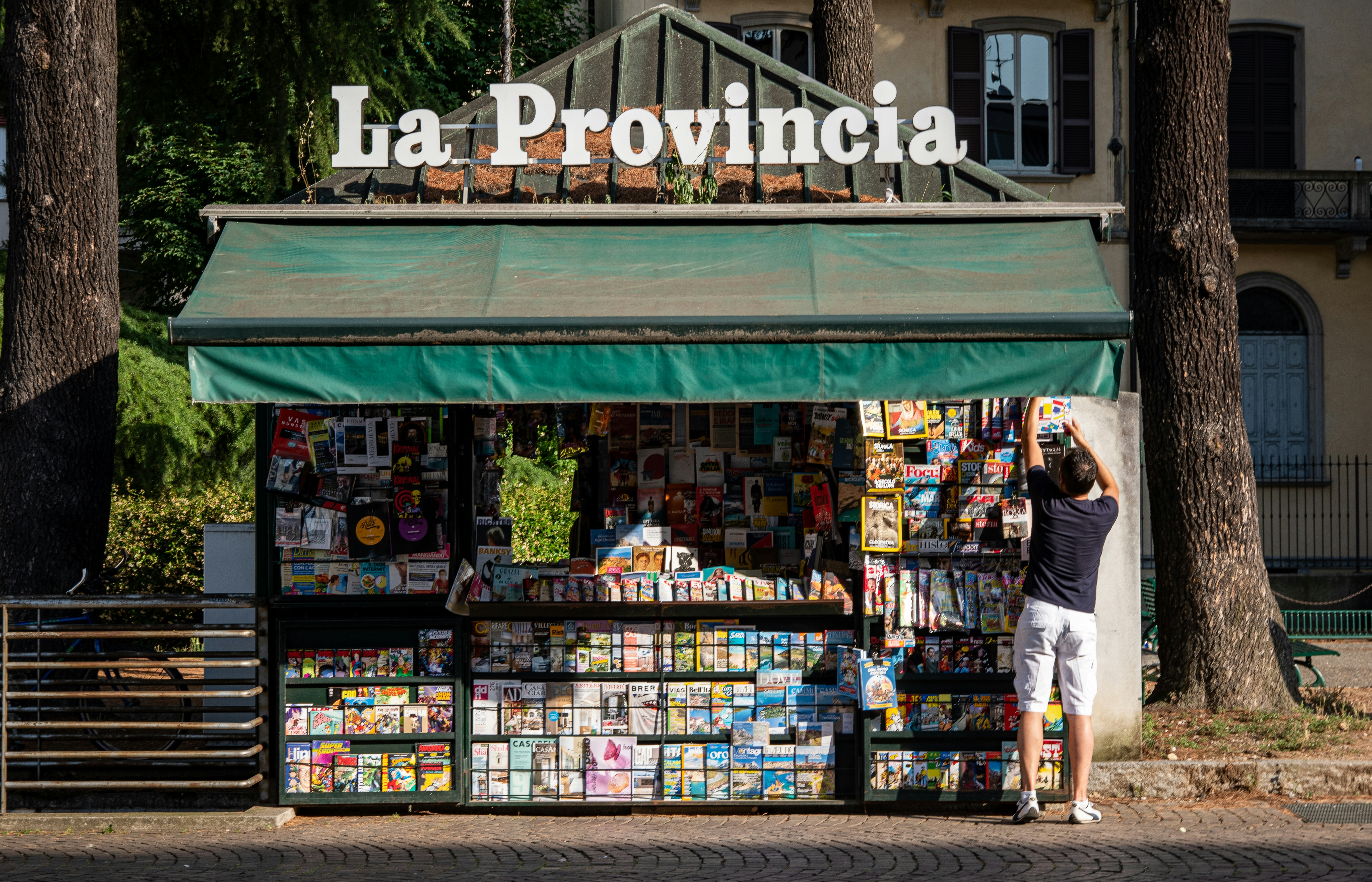 A bustling newsstand filled with colorful magazines and postcards, with a customer browsing the selection under a green awning.