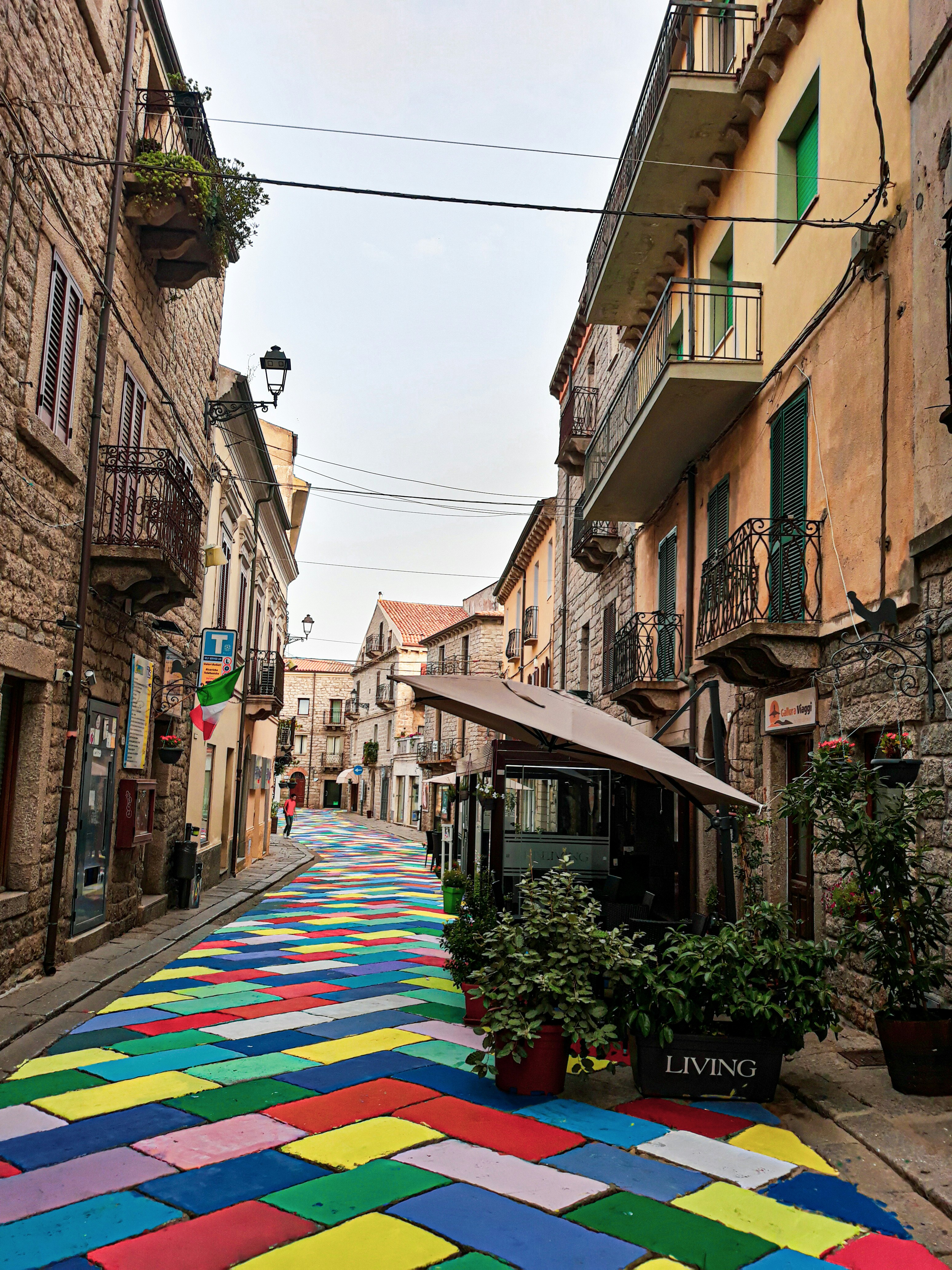 Colorful mosaic street in a historic European town with rainbow-hued paving, stone buildings, balconies, and a café awning along a pedestrian lane.