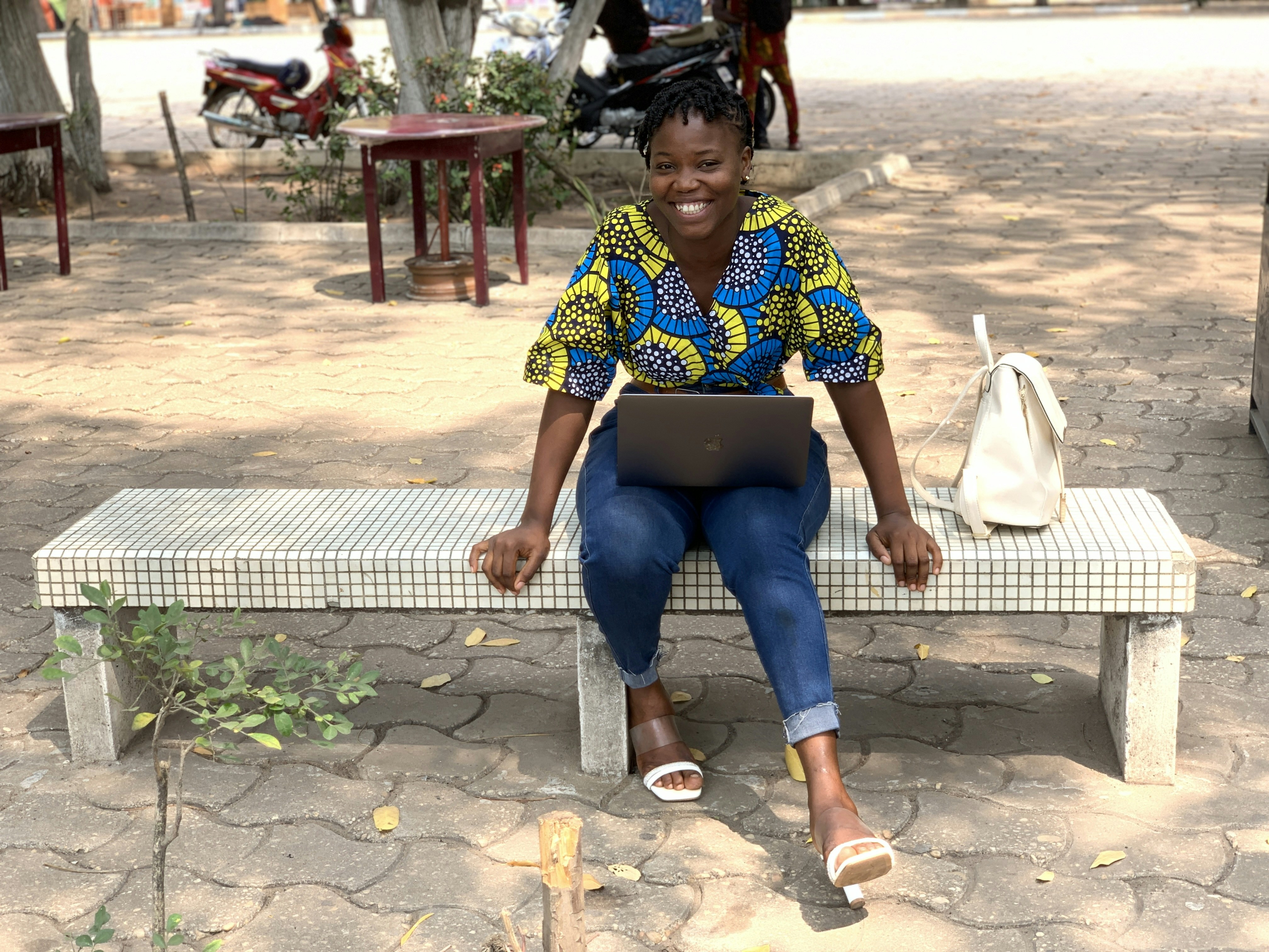 Woman smiling while seated on a bench, working on a laptop in a lively outdoor environment. Brightly colored attire contrasts with the serene background.