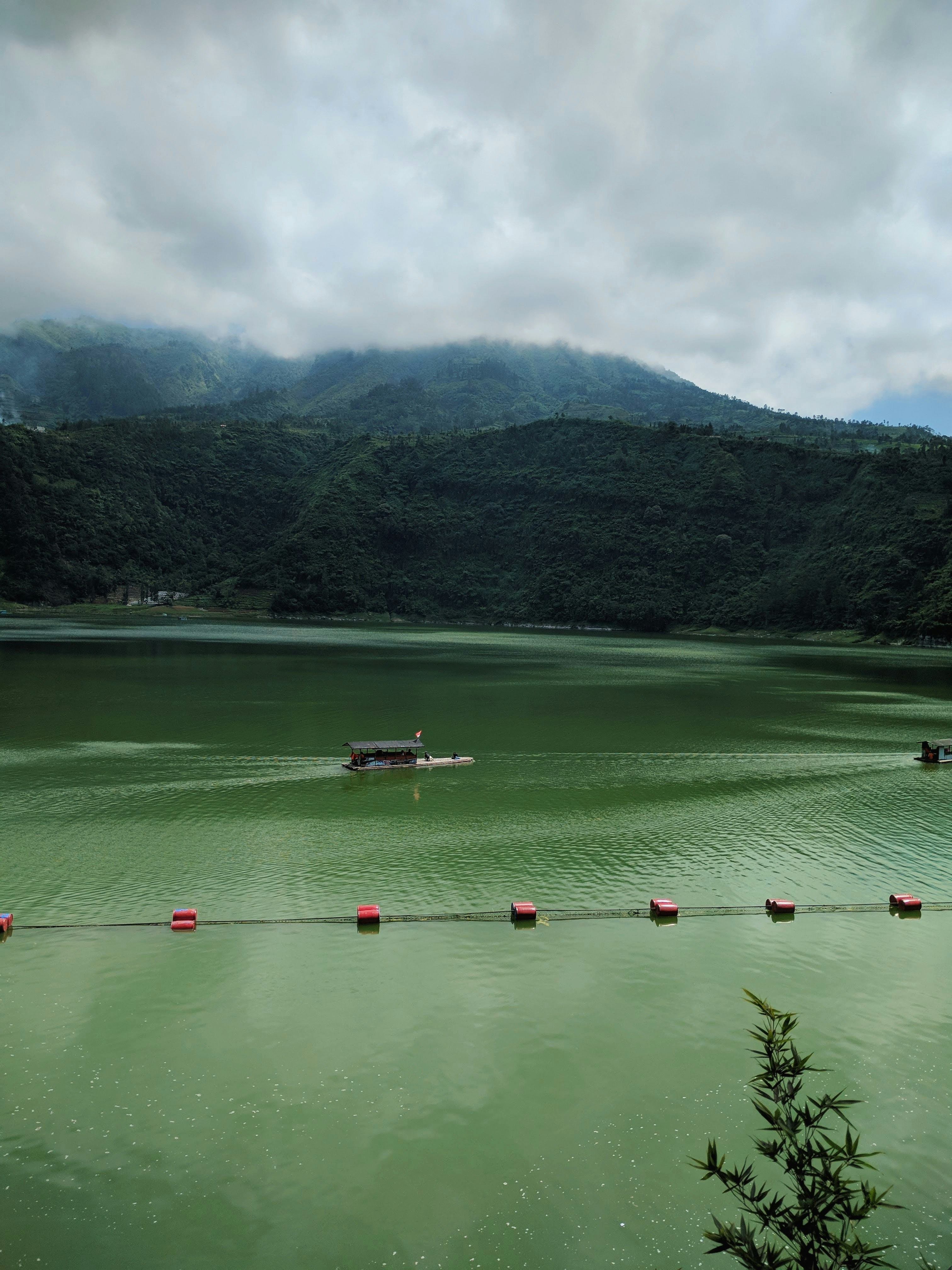 A tranquil boat glides across a verdant lake, surrounded by lush mountains and a cloudy sky. The scene captures the harmony of nature and human activity.