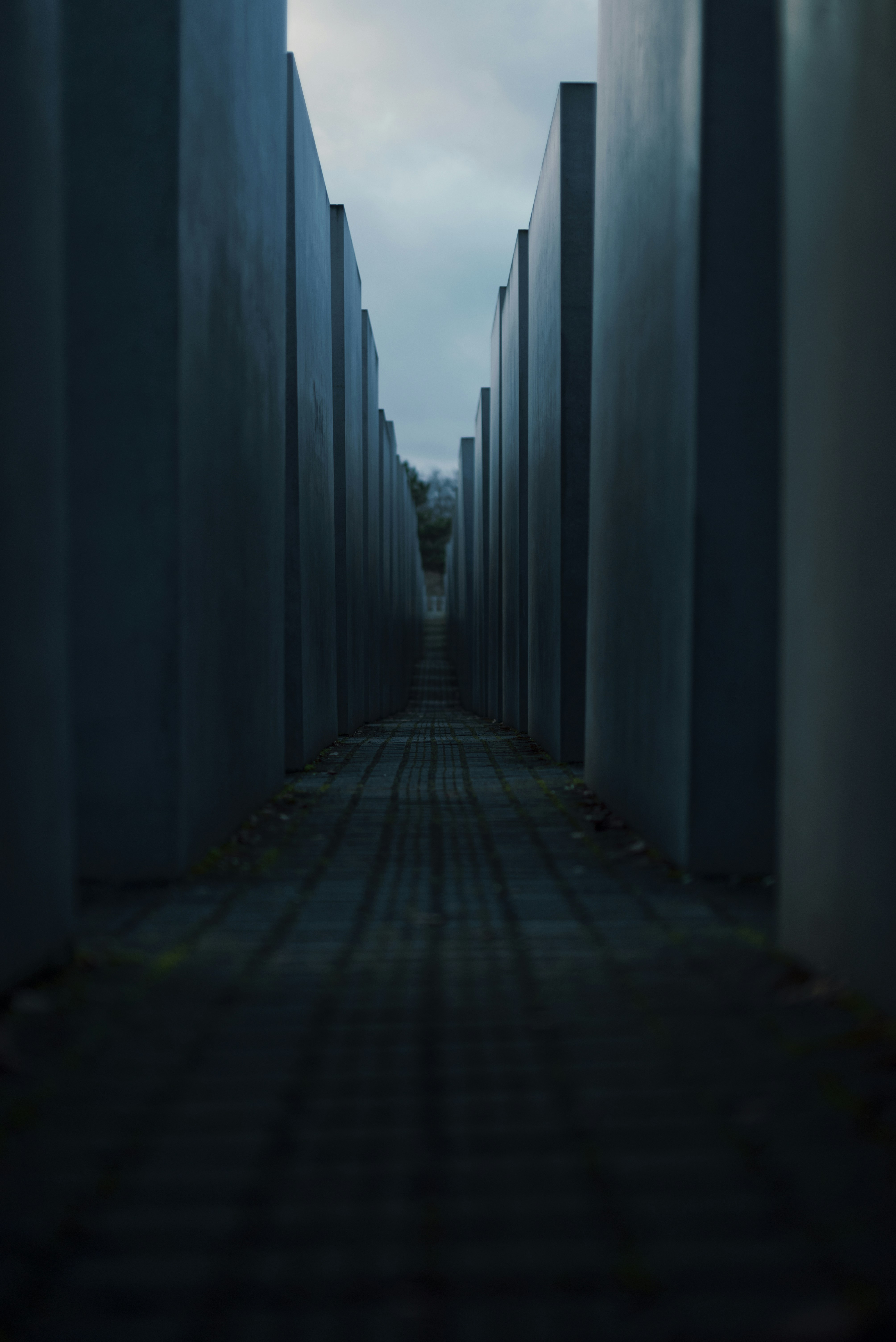 Narrow pathway flanked by tall, stark concrete slabs under a moody sky, evoking a sense of reflection and solemnity.